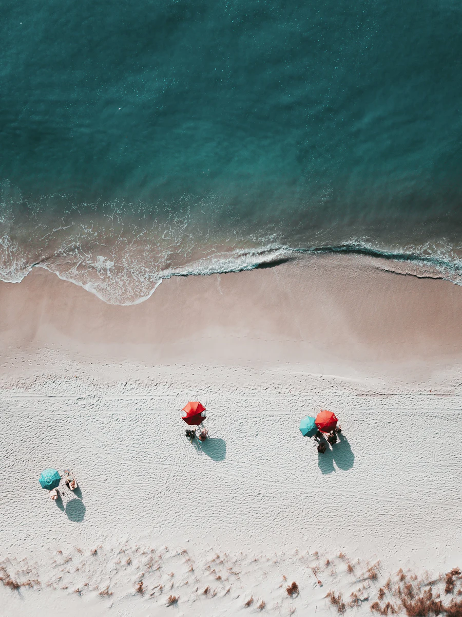 Aerial view of three colorful beach umbrellas on pale sand beside turquoise water and a curling line of surf. Long shadows stretch across the empty shoreline.
