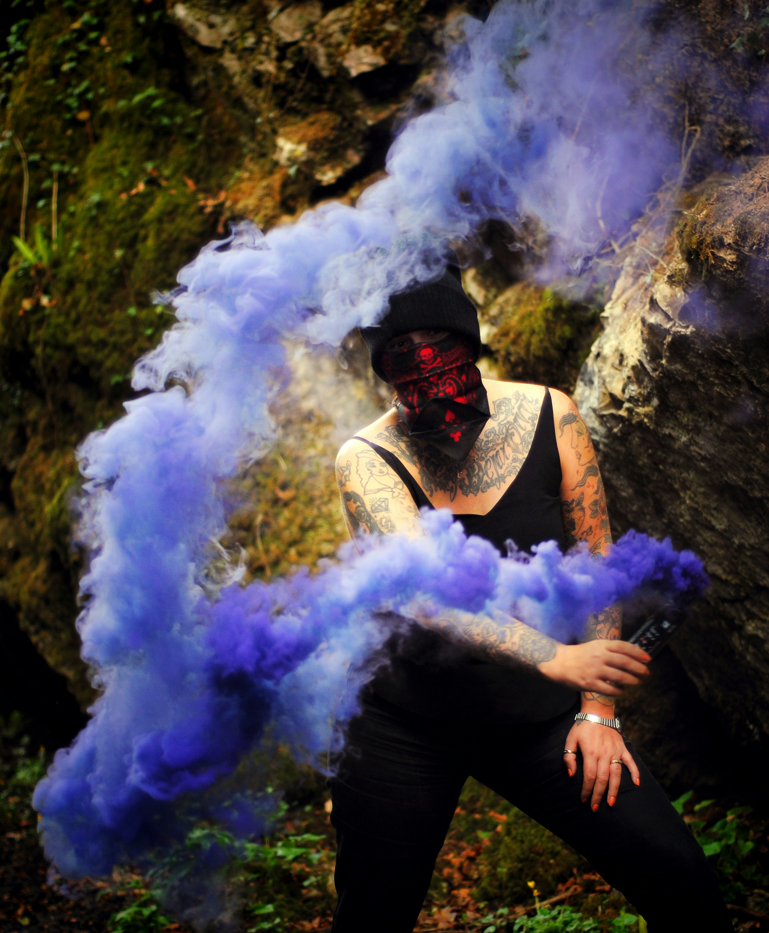 A figure shrouded in purple smoke, partially obscured by a natural backdrop, creates a striking contrast with their tattooed arm and black attire.