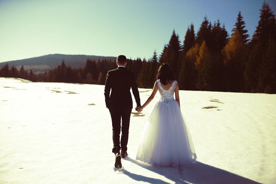 A bride and groom walk hand in hand across a bright snowy field with evergreen trees and mountains in the background. Their long shadows stretch behind them in the low sun.