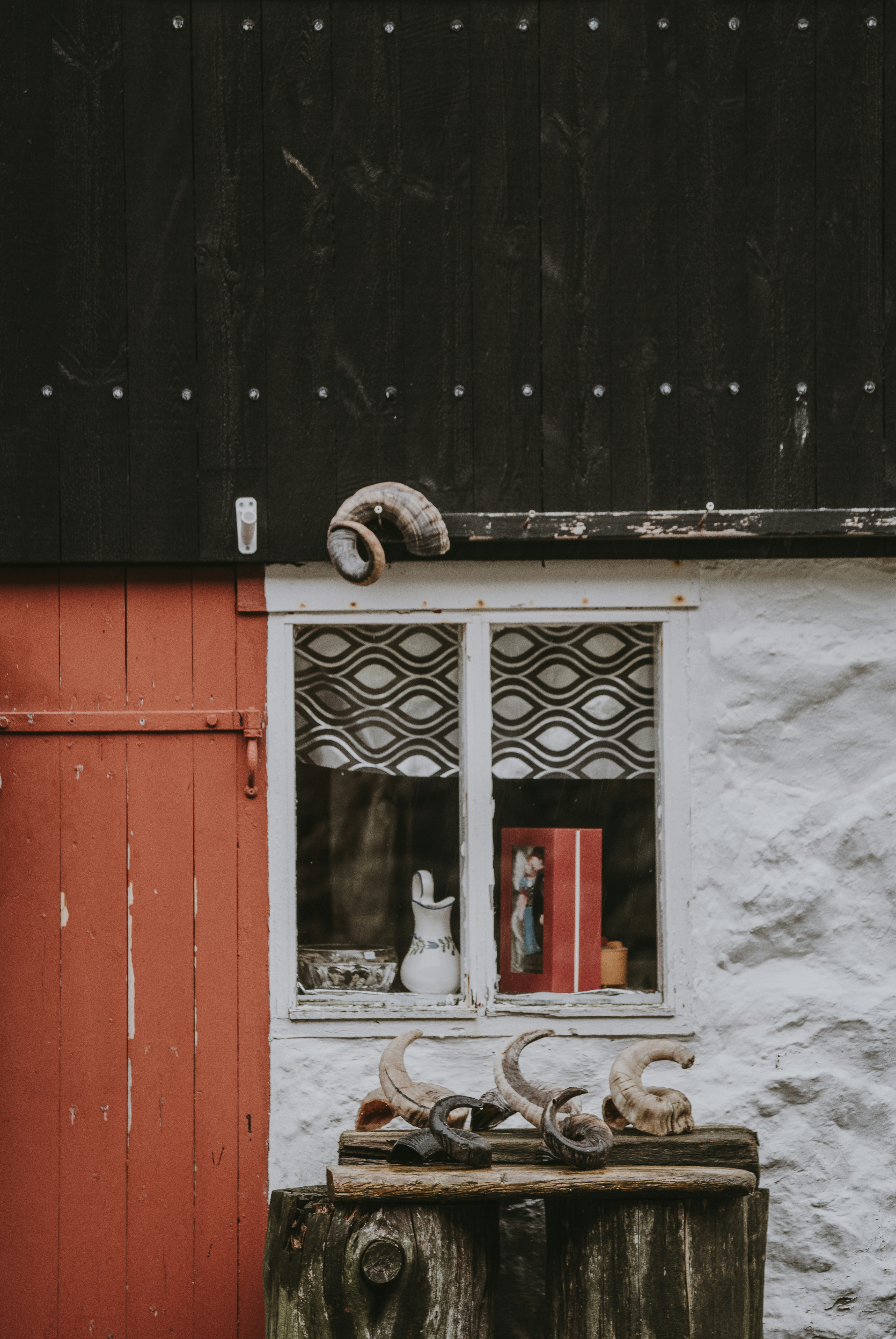 A quaint country cabin featuring a window adorned with decorative elements and rustic horns resting on a wooden table below. The contrasting colors of the red door and white wall enhance the cozy atmosphere.
