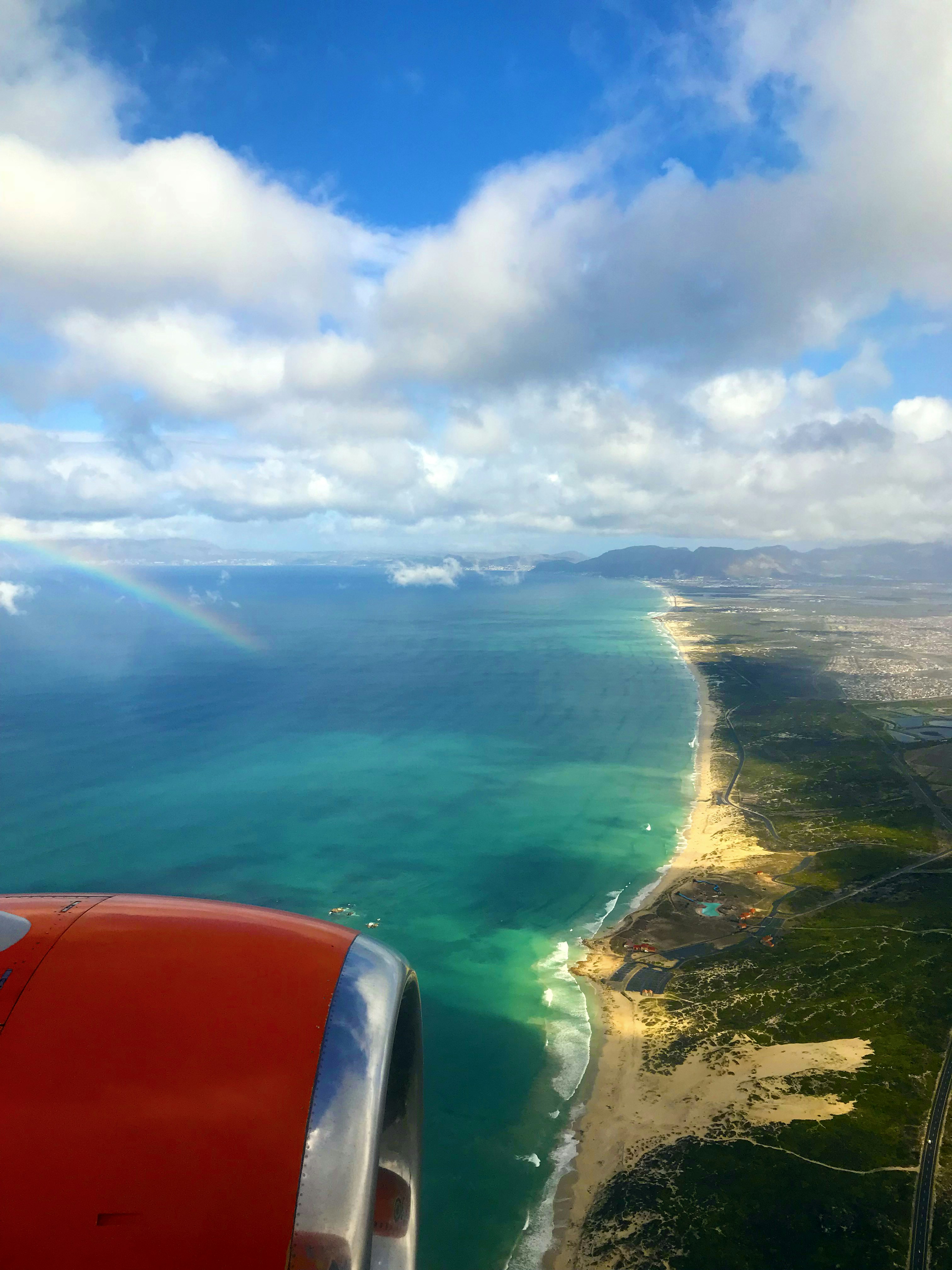 Aerial view of a turquoise coastline with a rainbow over the water and an orange airplane wing in the foreground.