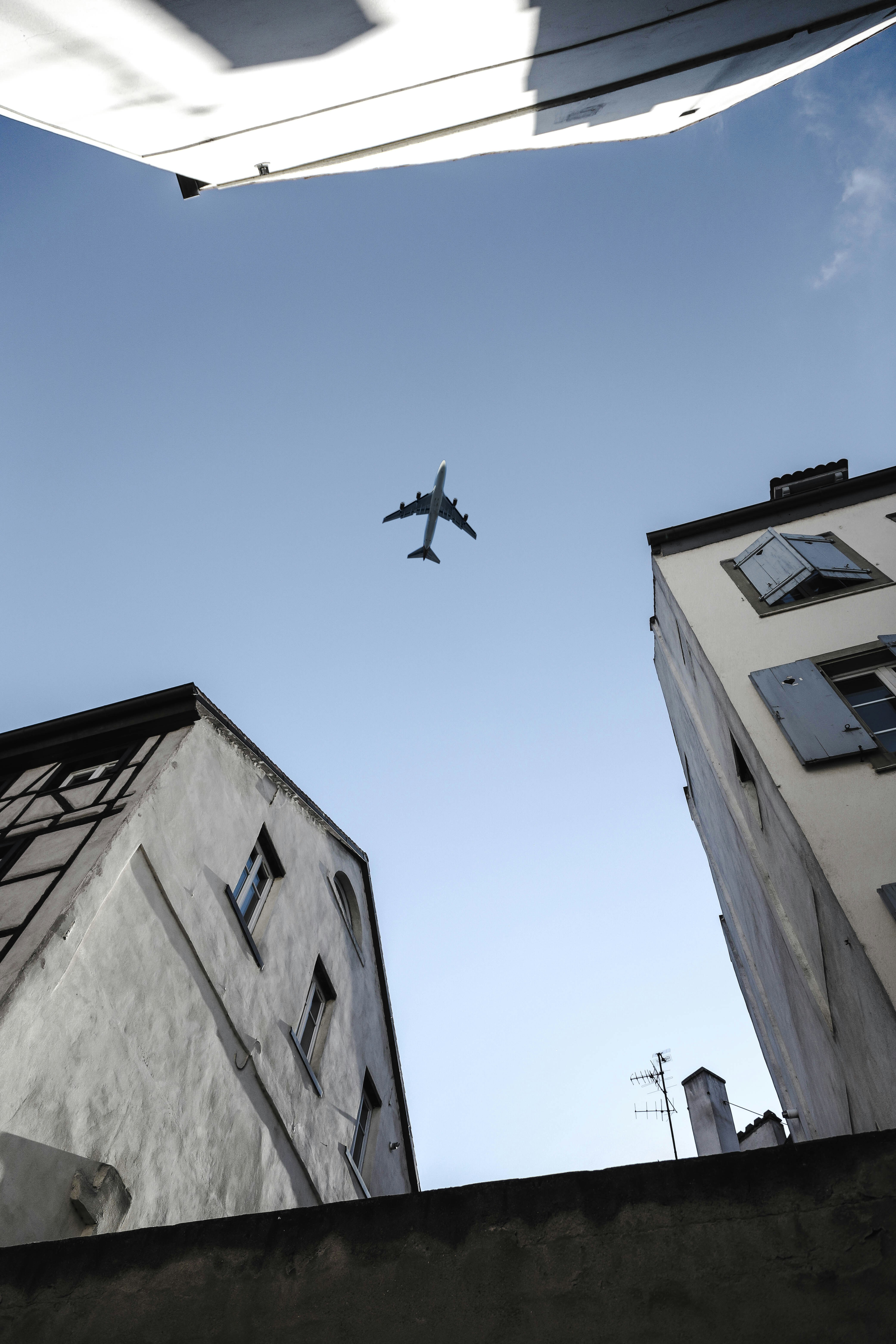 Passenger jet crosses the clear blue sky between tall, weathered buildings seen from a narrow street.