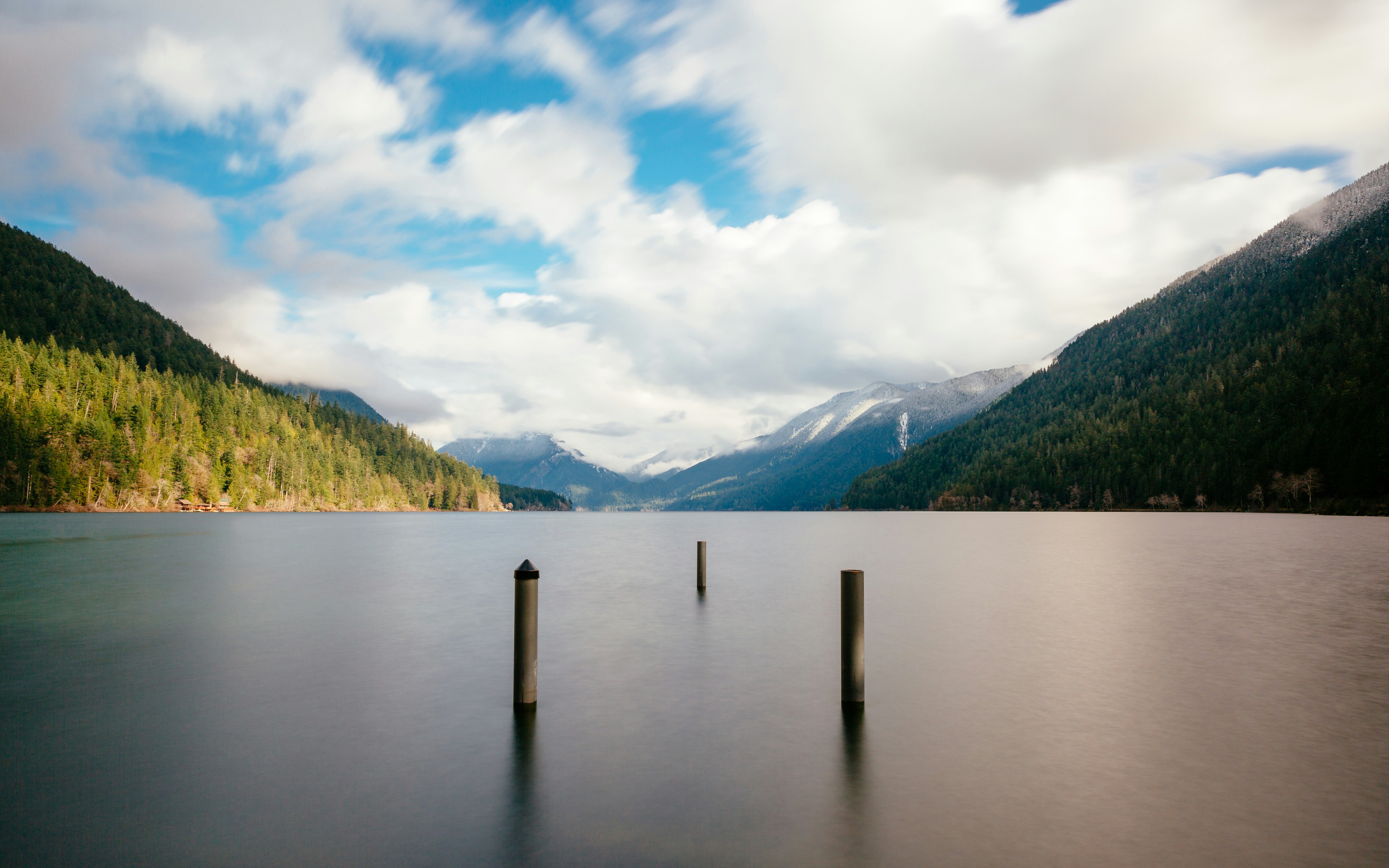 Serene lake with mountain backdrop and three wooden posts emerging from the water under a partly cloudy sky.