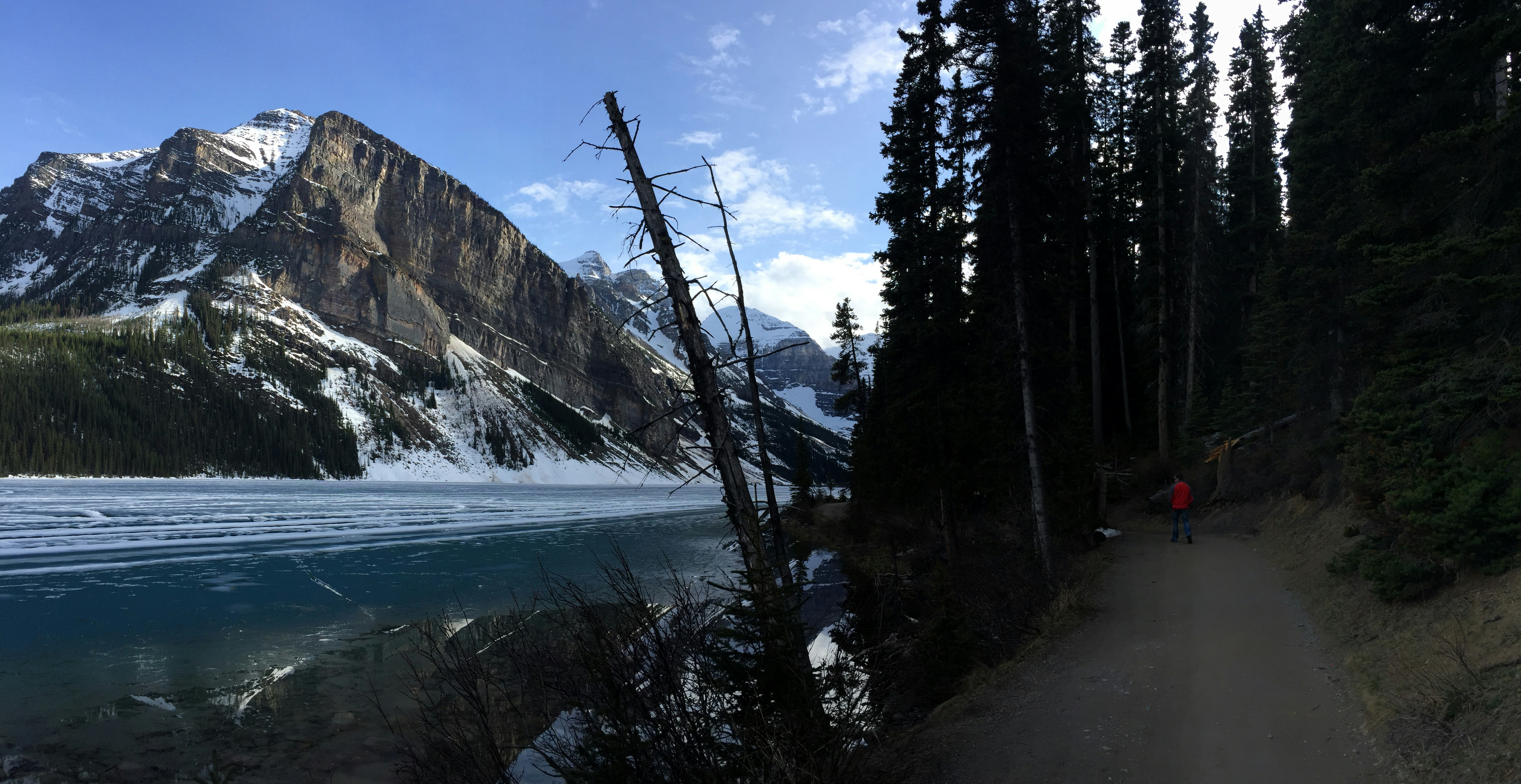 Alpine trail runs along a partially frozen lake beneath snow-dusted peaks, with a lone hiker in a red jacket among tall evergreen trees.
