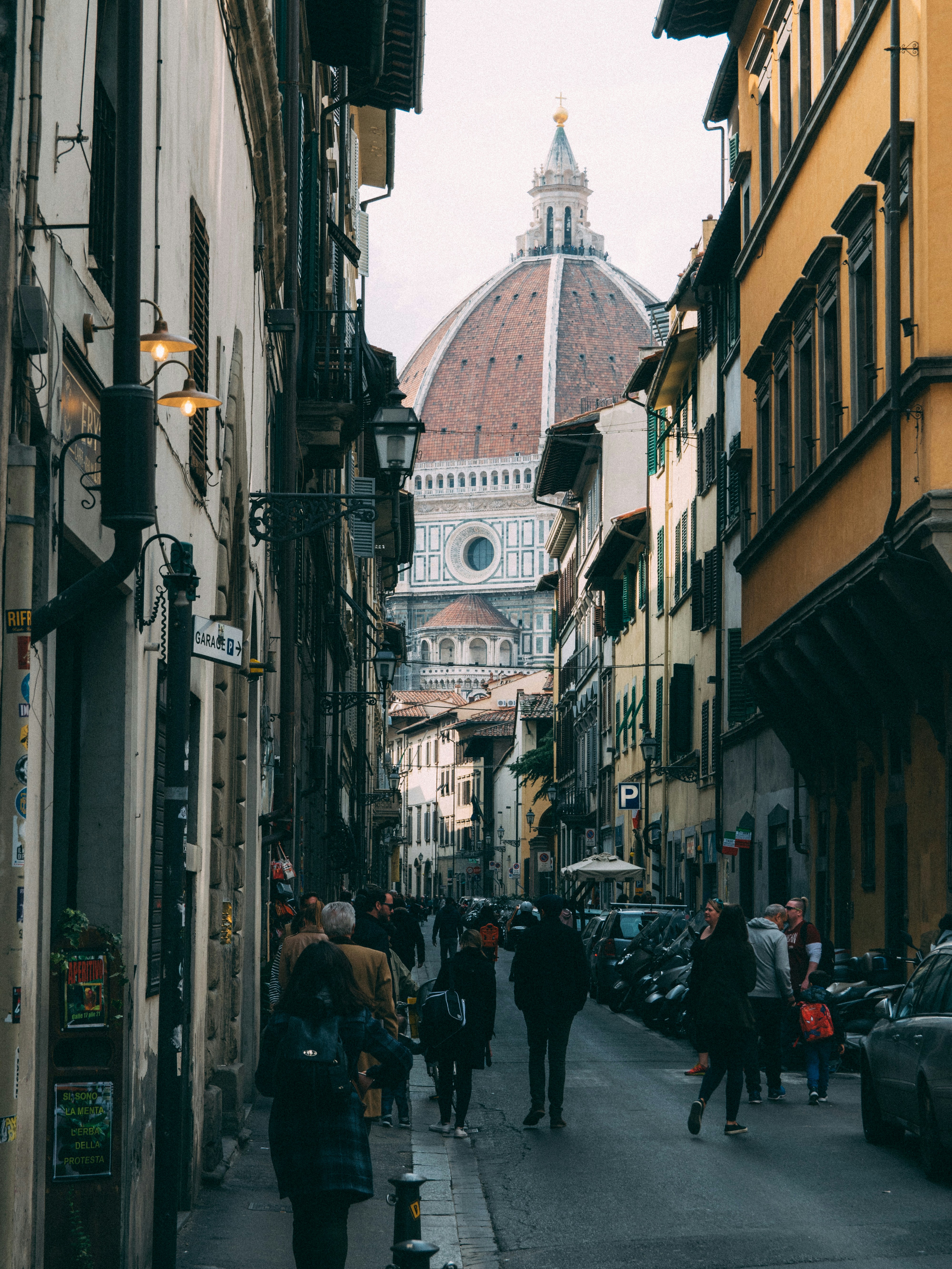 Narrow street in Florence leading towards the iconic dome of the Cathedral of Santa Maria del Fiore, bustling with pedestrians and lined with charming buildings.