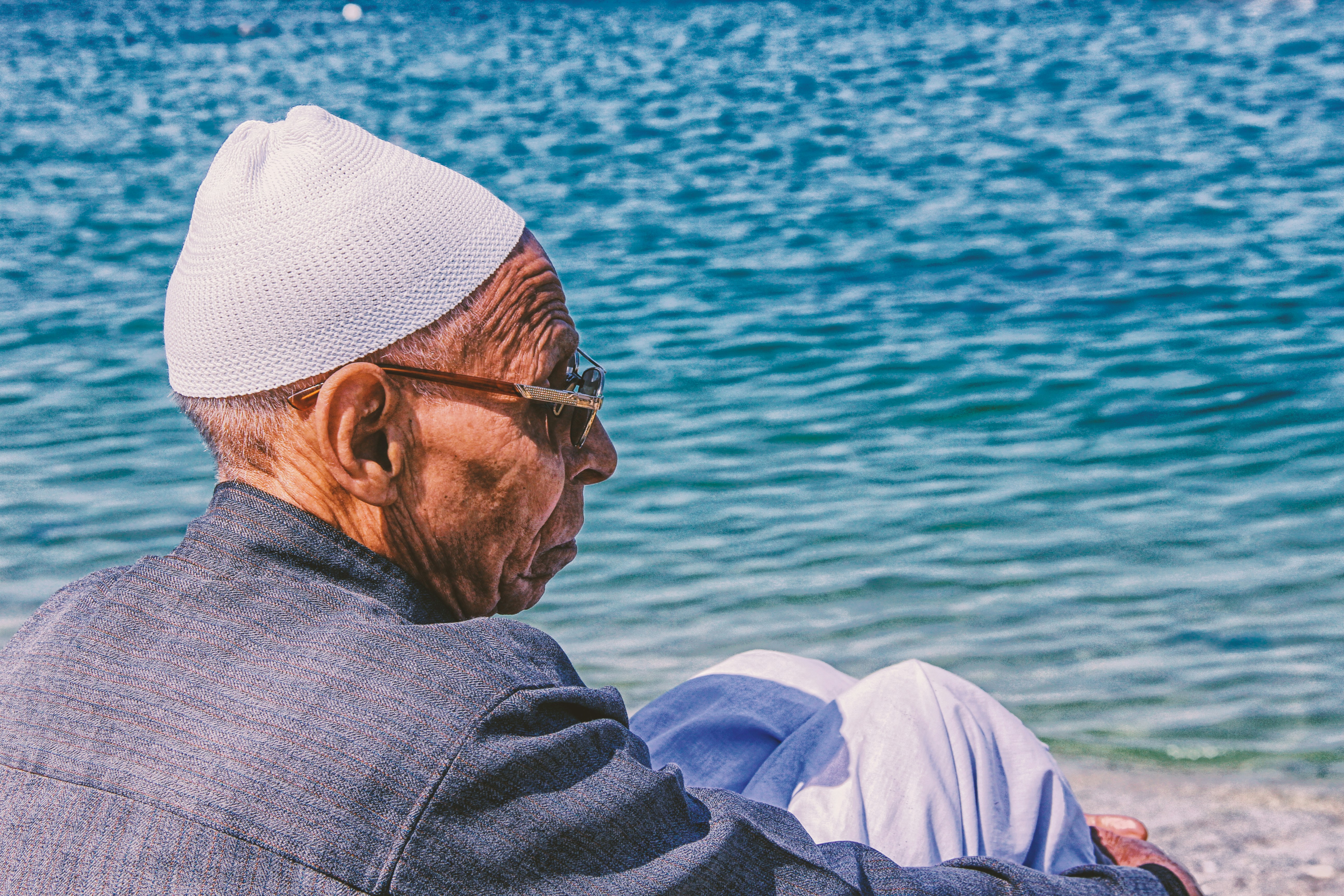 Elderly man in a white cap sitting on a beach facing the ocean.