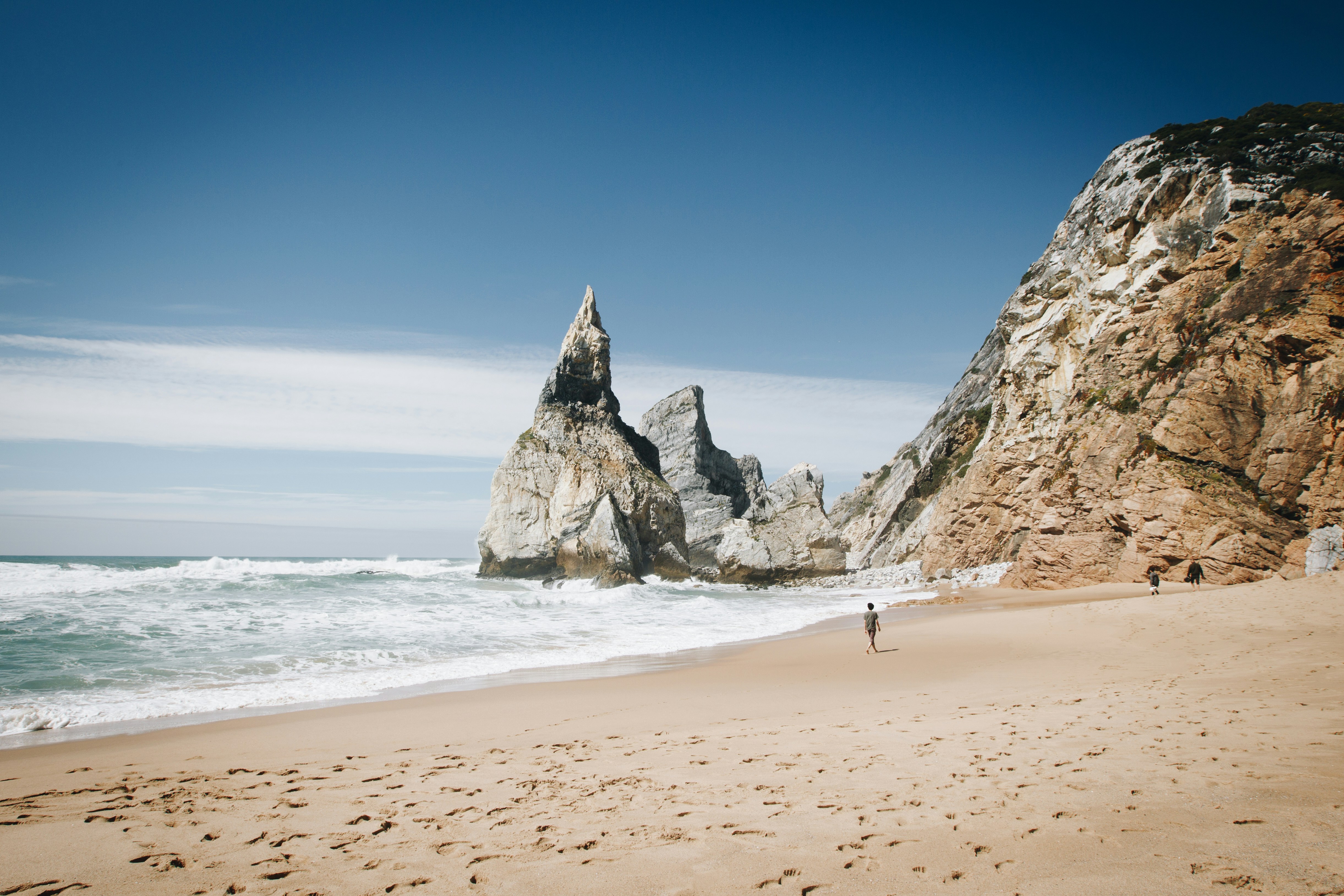 Person walking along a sandy beach near towering coastal rock formations under a clear blue sky.
