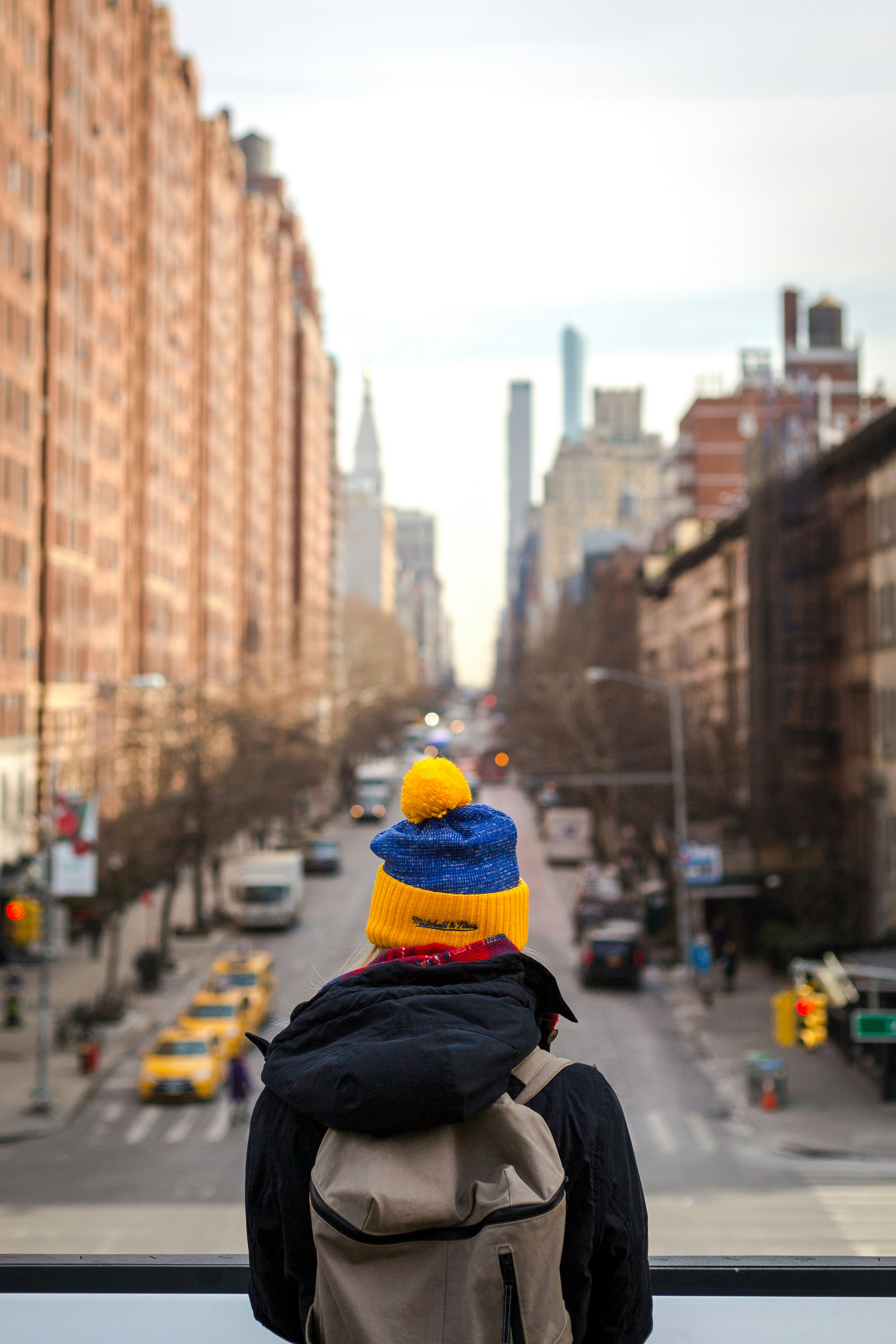 Individual wearing a colorful beanie gazes down a bustling city street, framed by towering buildings and vibrant yellow taxis. The scene captures the essence of urban exploration.