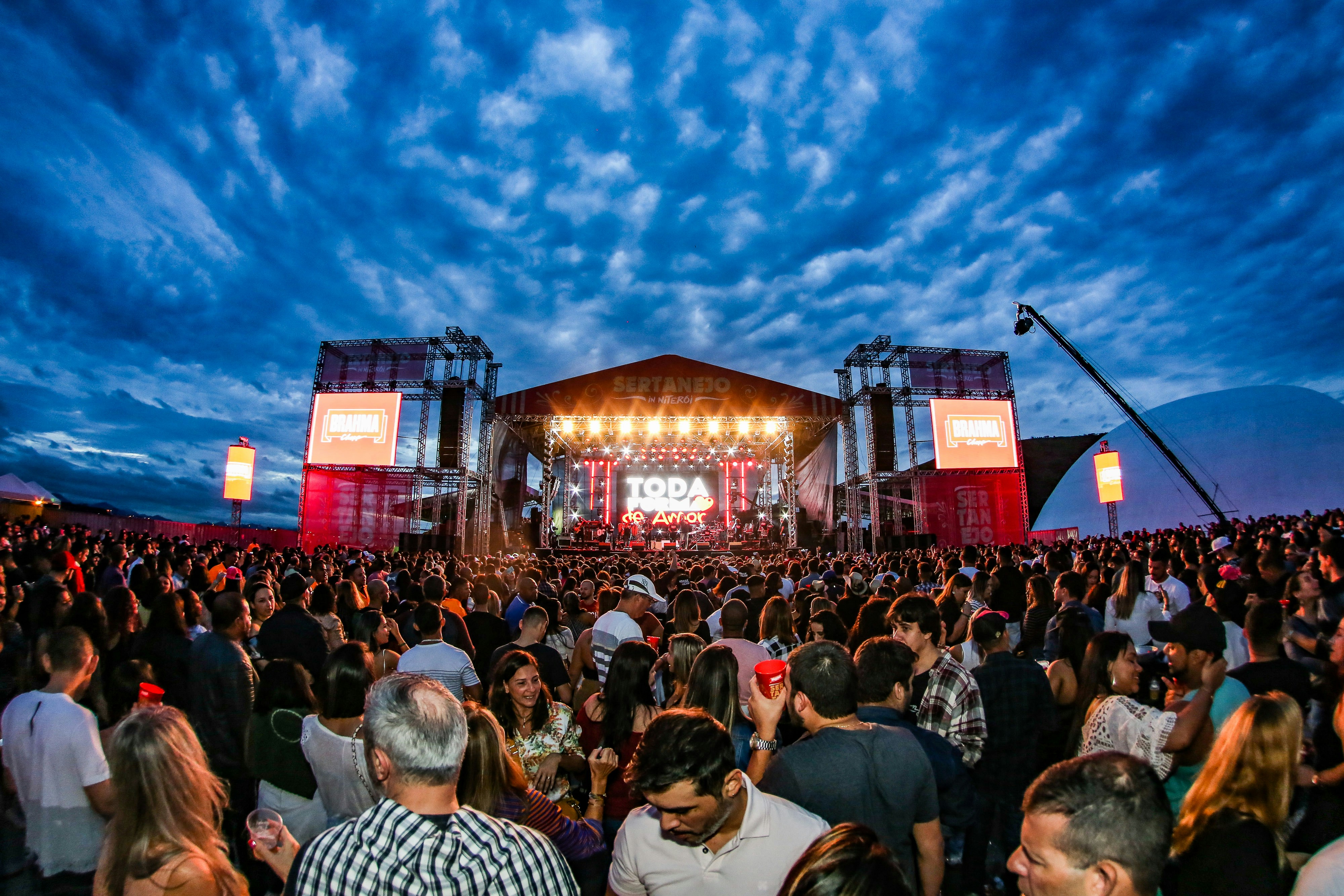 Large crowd gathered at an outdoor concert with a vibrant stage and dramatic evening clouds.
