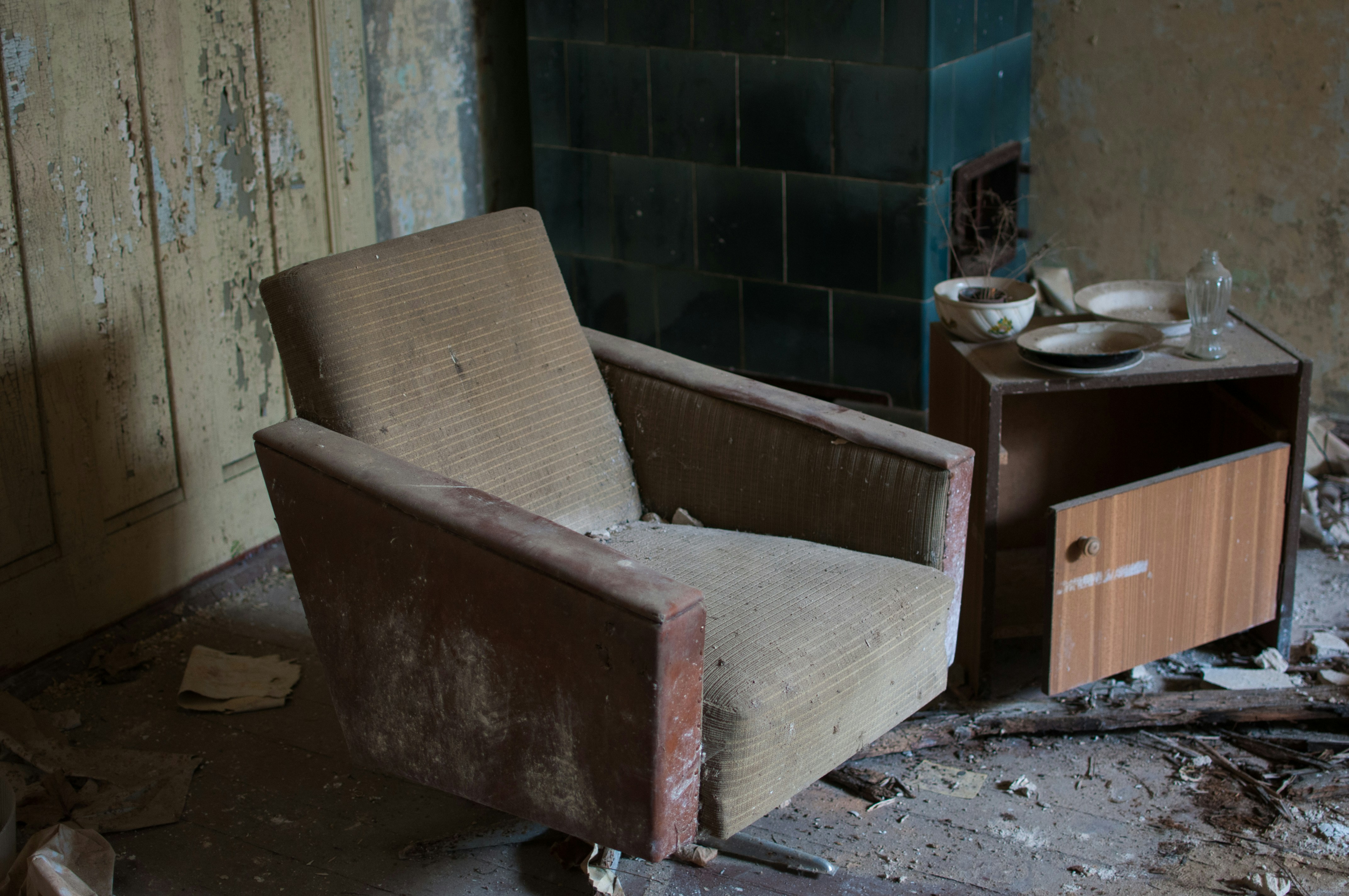 Abandoned armchair beside a wooden nightstand, surrounded by dust and debris in a neglected room. A few dishes rest on the table, hinting at past life.