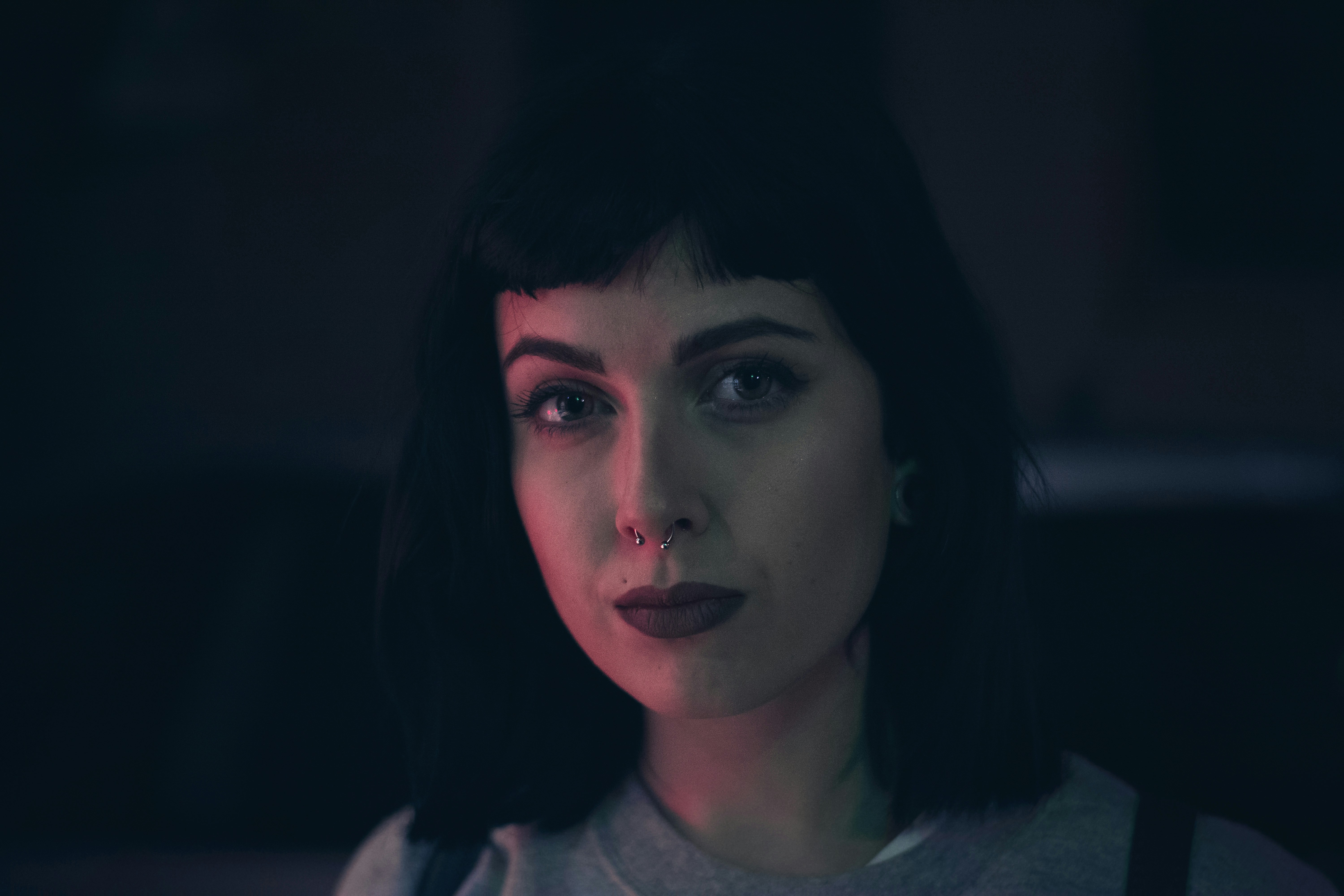 A young woman with dark hair and bold makeup gazes intently, illuminated by soft neon light in an urban setting.