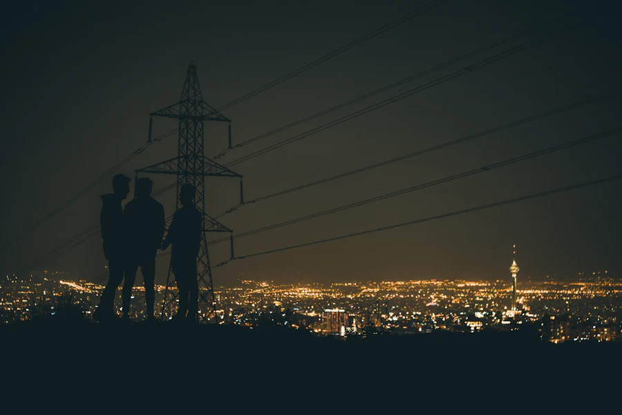 Three silhouetted figures stand on a ridge beside a power pylon, overlooking a glowing city skyline at night. Power lines stretch across the dark sky toward a lit tower in the distance.