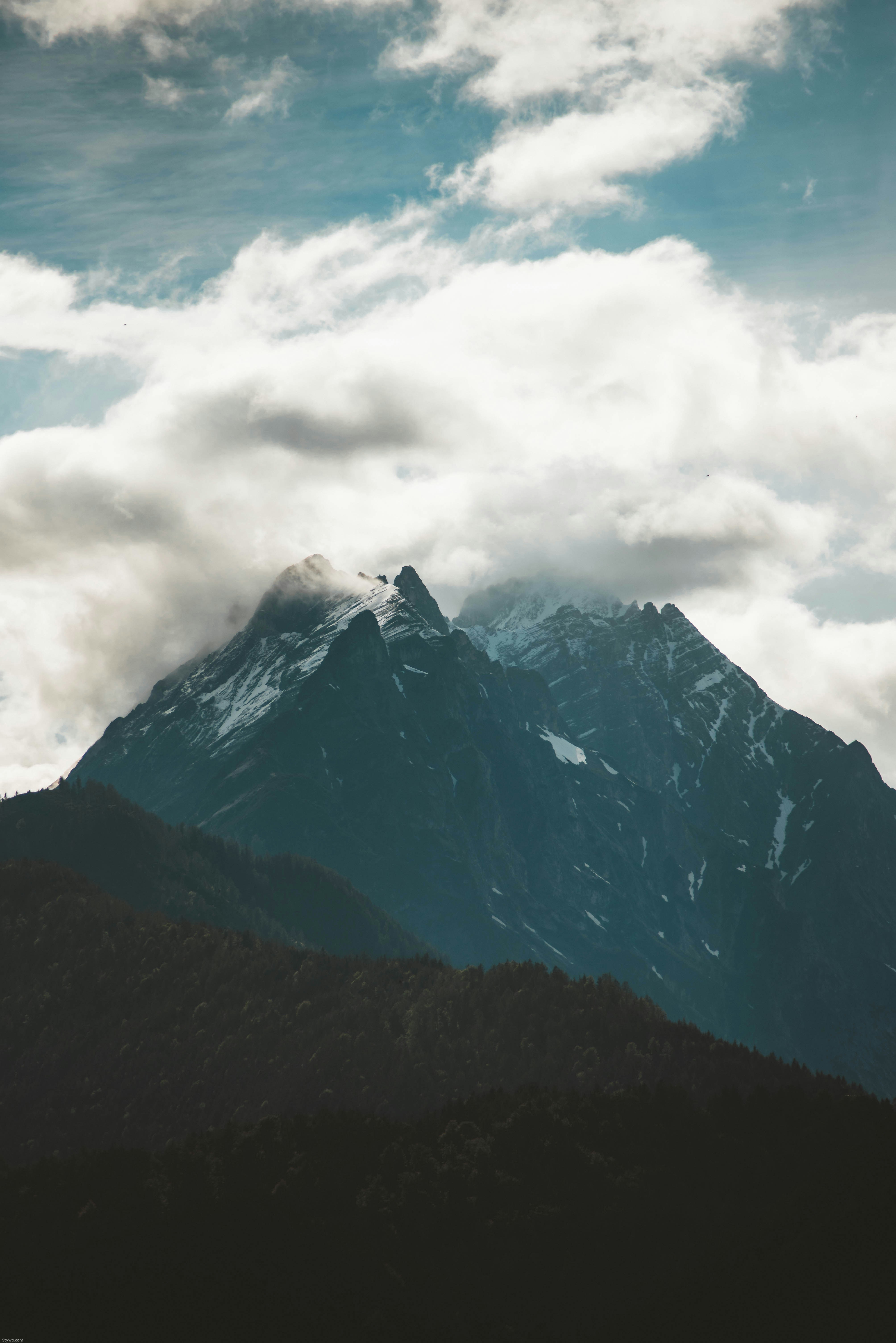 Snow-capped mountain peaks shrouded in clouds, rising dramatically against a moody sky. The interplay of light and shadow enhances the rugged terrain.
