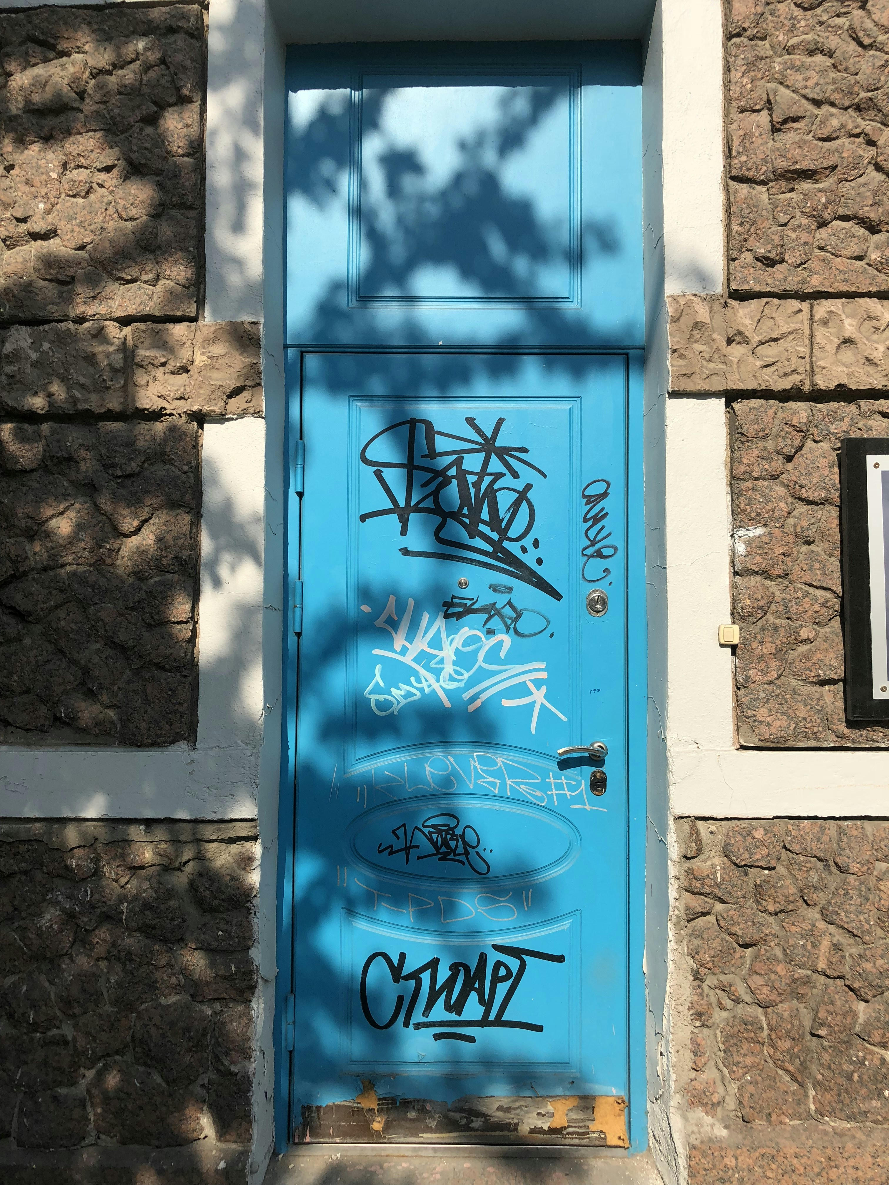 Vibrant blue door adorned with various graffiti tags, framed by textured stone walls. The interplay of light and shadow adds depth to the urban scene.