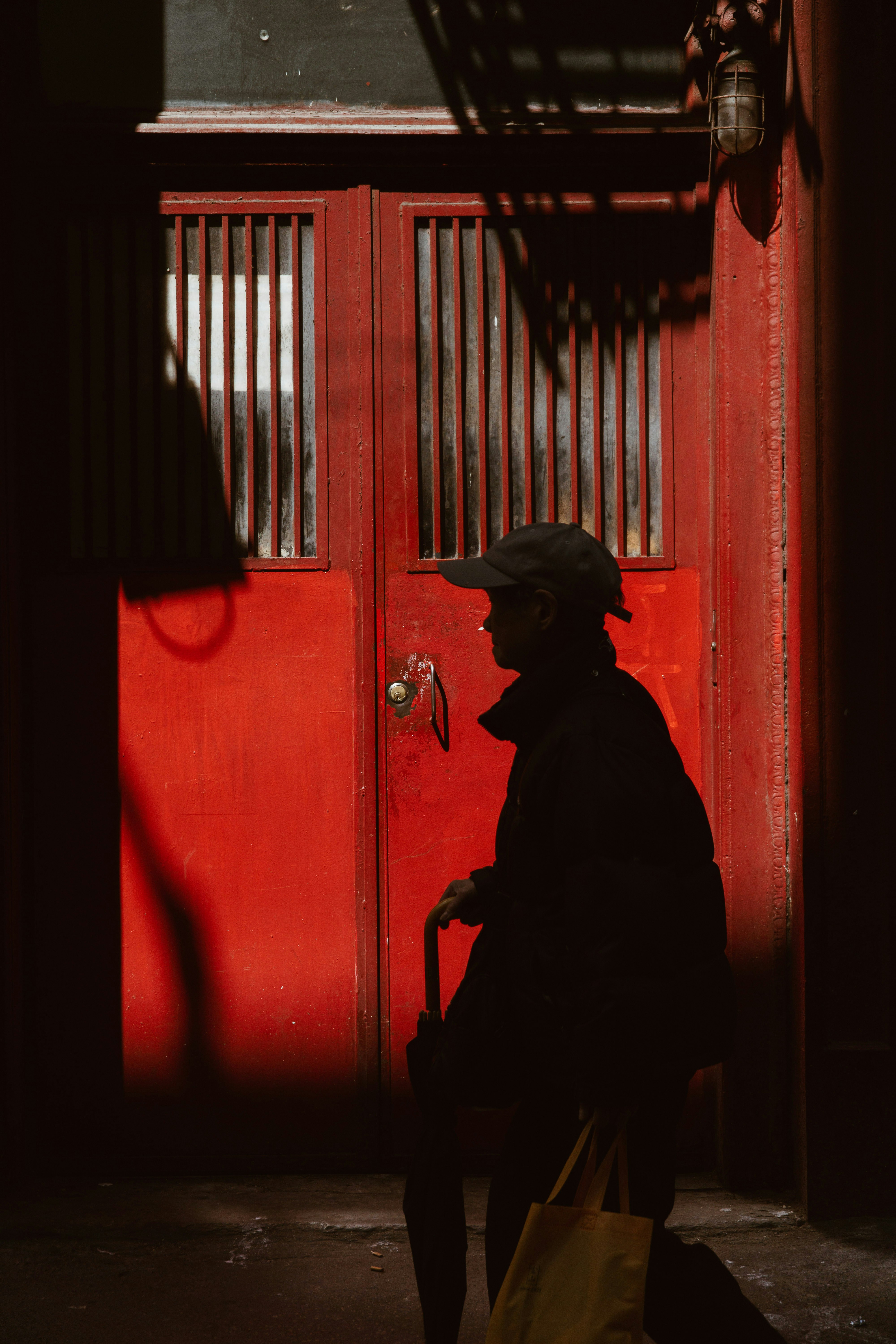 Silhouette of a person walking past a vibrant red door, highlighted by contrasting shadows. The scene captures a moment of urban life infused with rich colors.