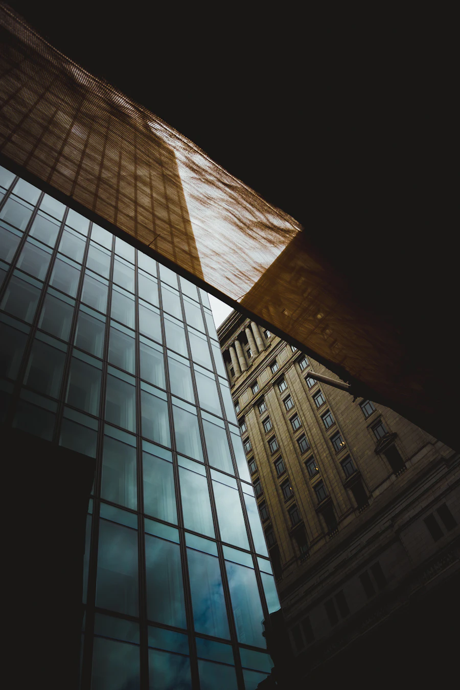 Low-angle view of a modern glass building beside an ornate stone facade, framed by a dark canopy overhead. Reflections and geometric lines create a moody urban scene.
