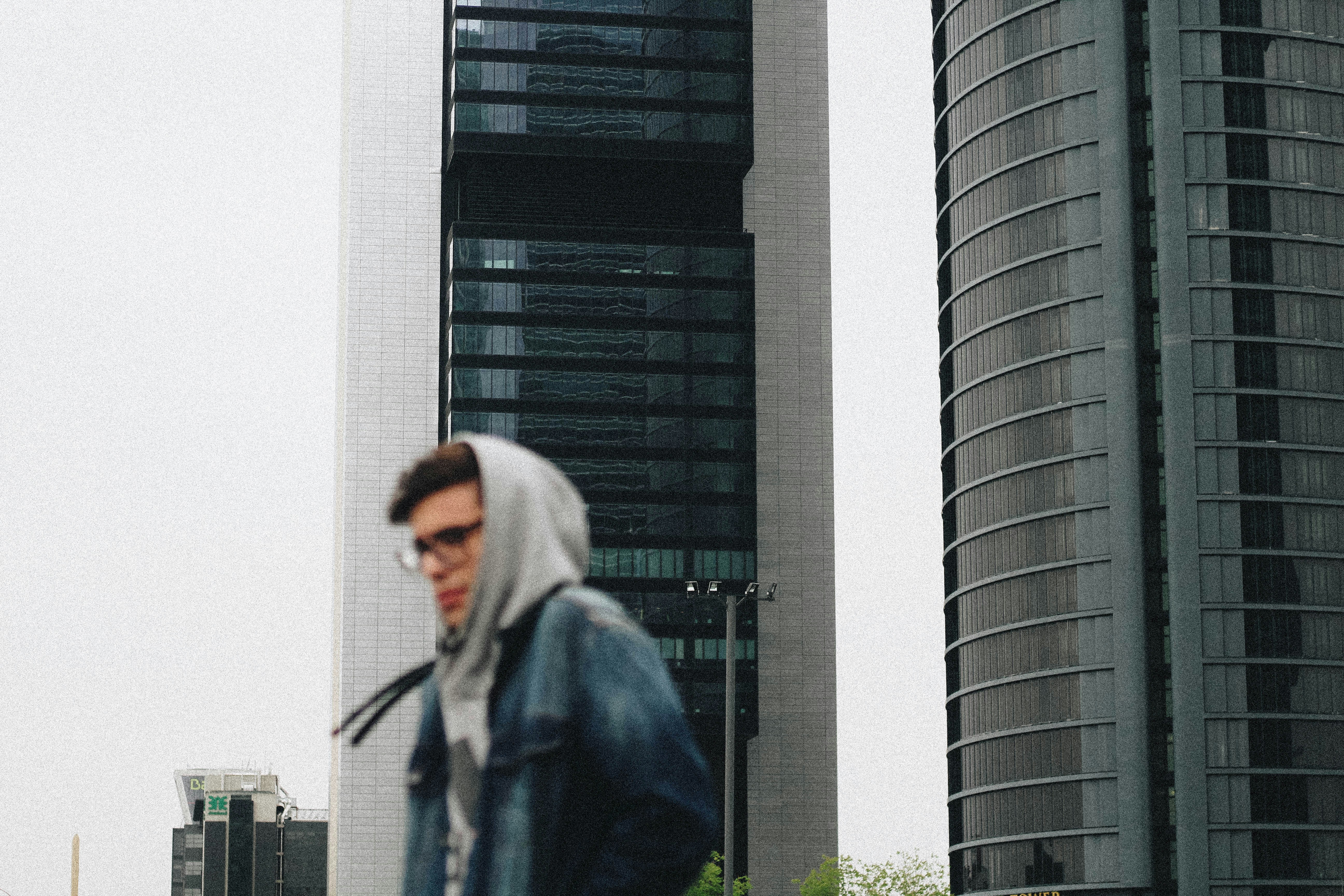 Person in a hoodie walking past modern skyscrapers under an overcast sky.