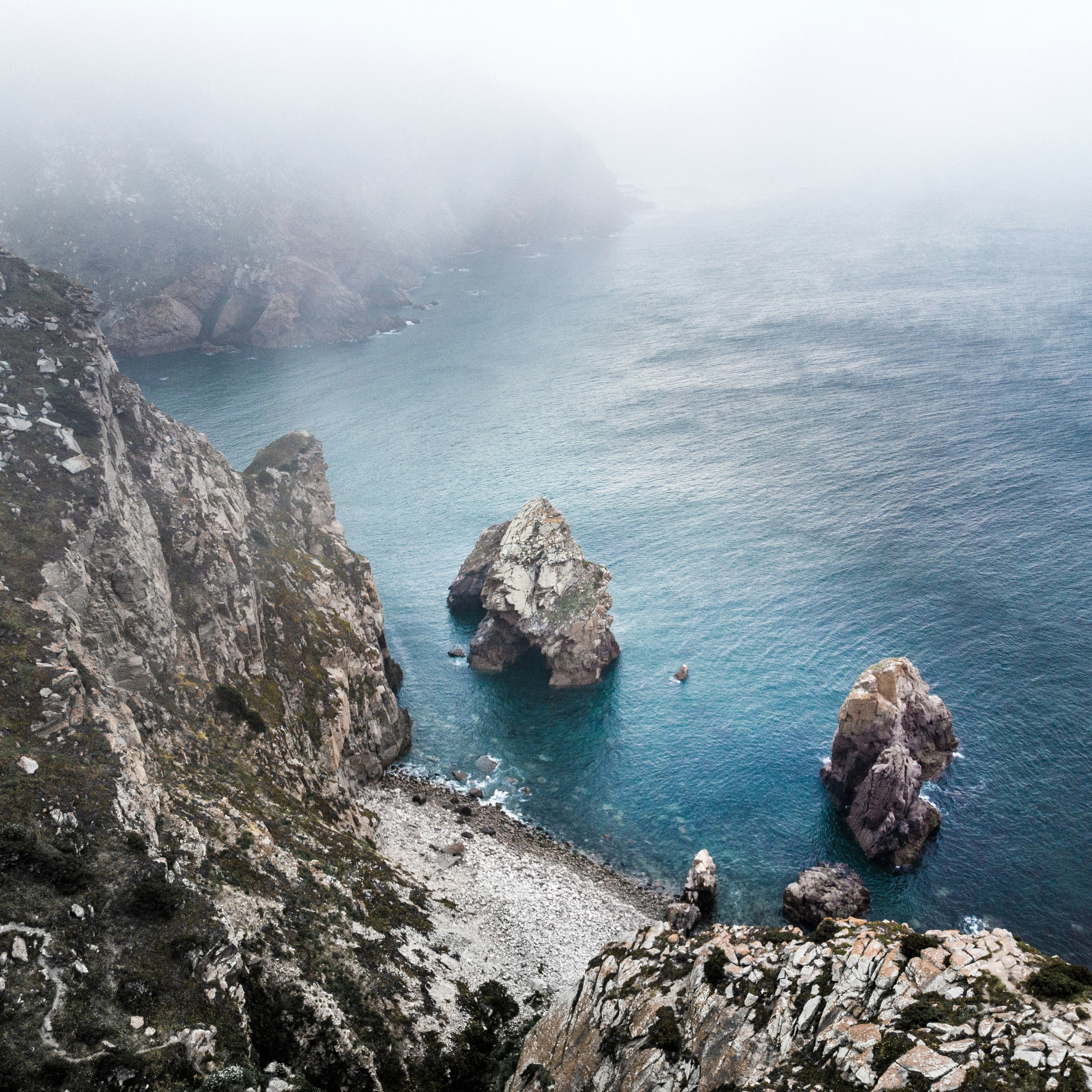 Foggy coastline revealing rugged cliffs and serene waters, with prominent rock formations jutting from the sea. A tranquil scene of nature's beauty.