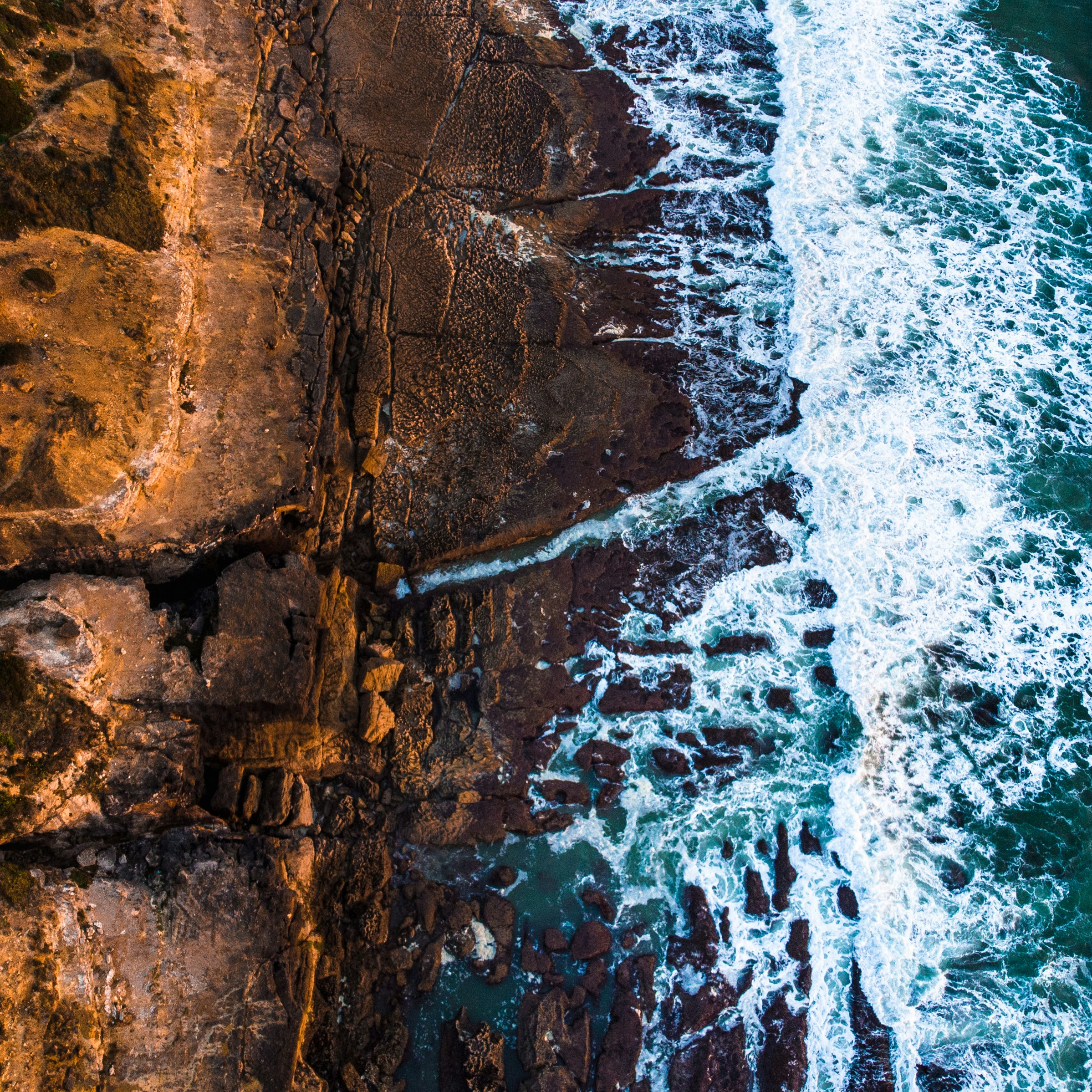 Aerial photograph of a rugged cliffline on the left meeting turquoise surf on the right, with sunlit rock and foamy waves creating a stark contrast.