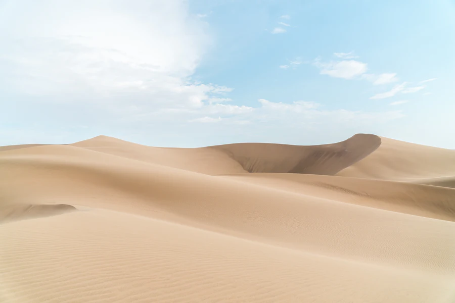 Soft sand dunes under a pale blue sky with wispy clouds, creating smooth curves and layered ridgelines. A dark hollow in the central dune adds contrast and depth.