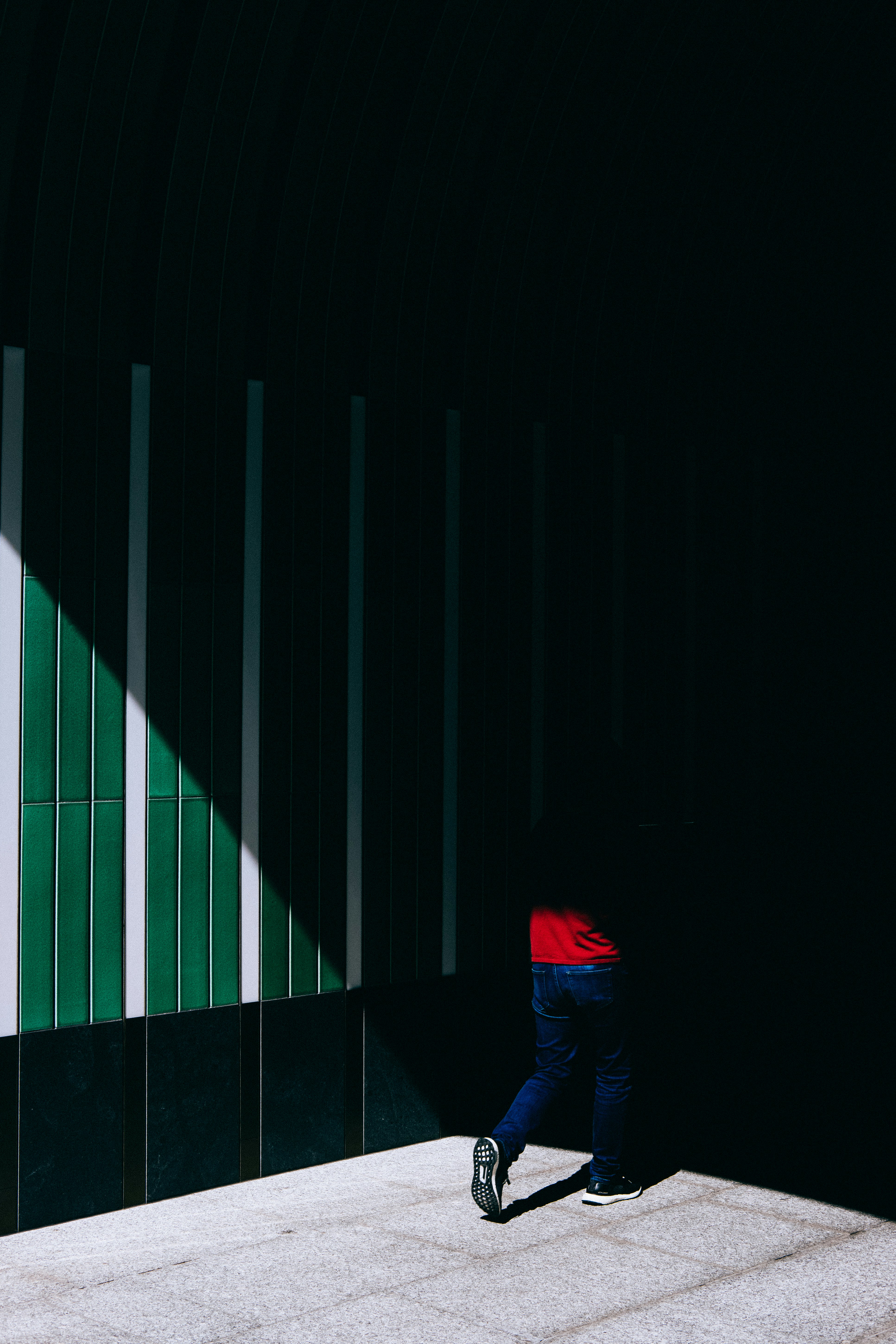 A figure in a red shirt walks along a wall of green tiles, contrasting sharply against deep shadows. The interplay of light and dark creates a dynamic urban scene.