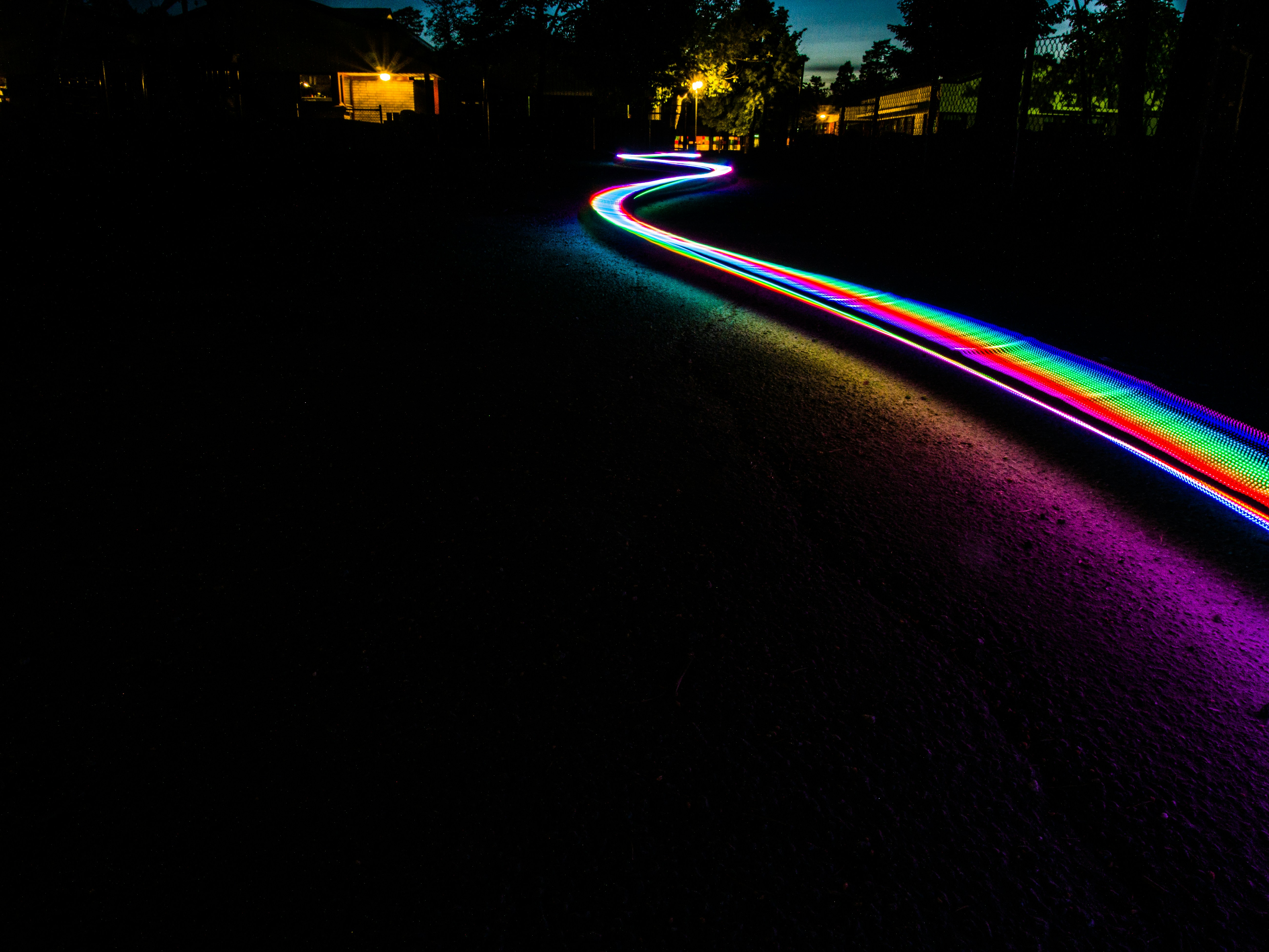 Long-exposure night shot of a neon rainbow light trail winding across the dark ground, with distant houses softly lit in the background.