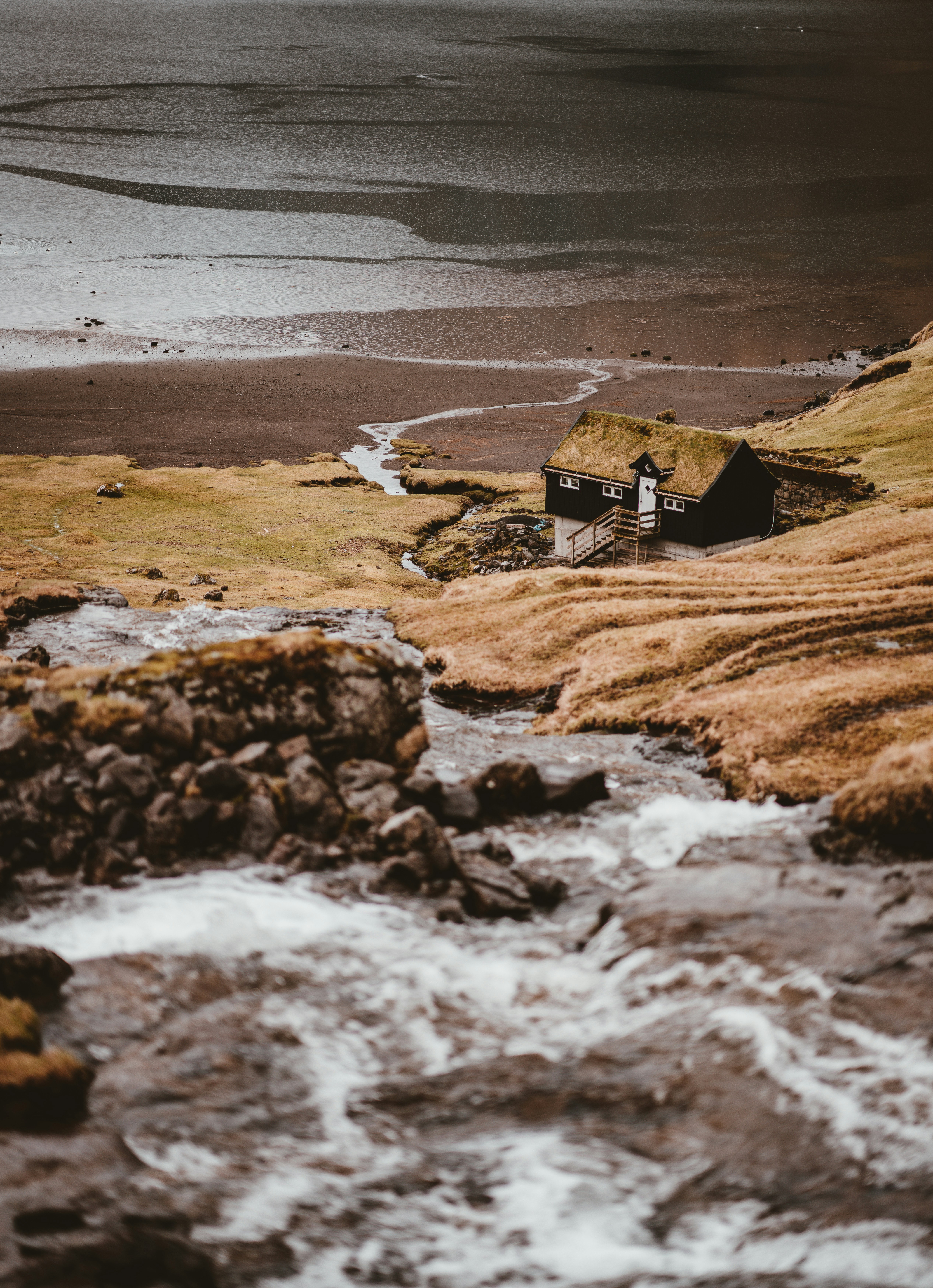 A quaint, thatched-roof house nestled beside a gentle stream, surrounded by lush green hills and a serene shoreline. The landscape reflects a harmonious blend of nature and human habitation.