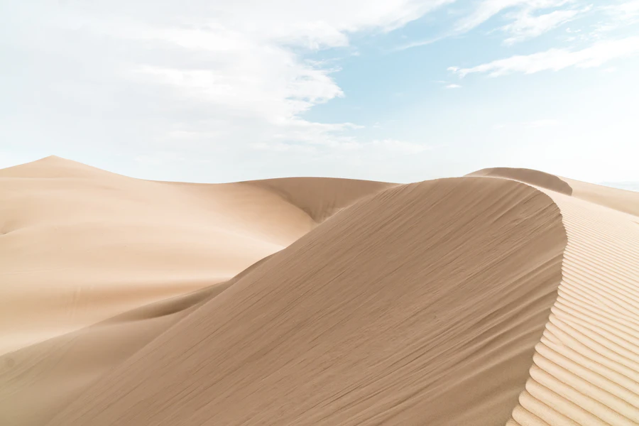 Wind-sculpted sand dunes curve across a bright desert landscape beneath a soft blue sky with wispy clouds. Fine ripples and layered slopes create a flowing, minimal scene.