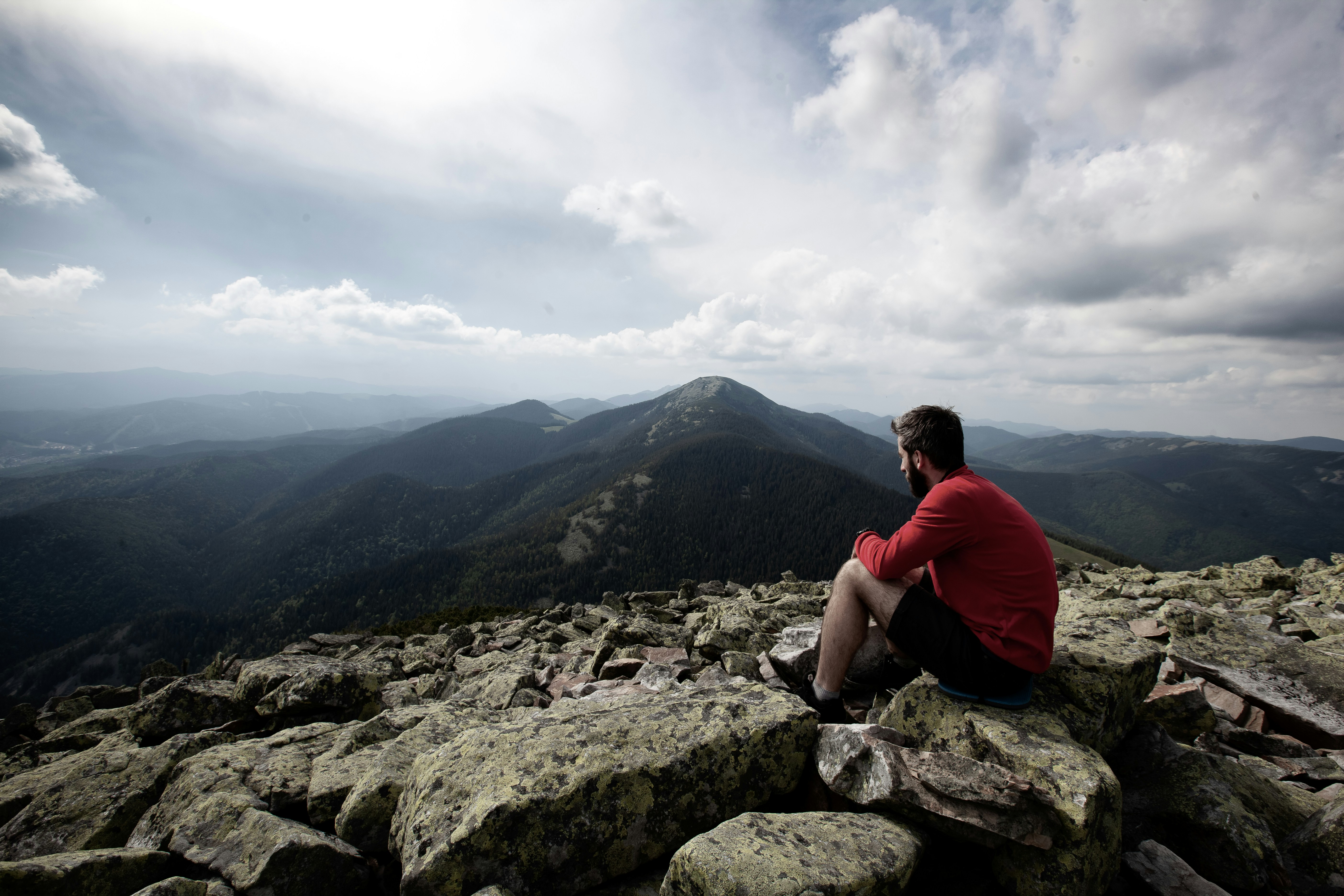 Person in red jacket sitting on rocky terrain, gazing at distant mountain under a cloudy sky.