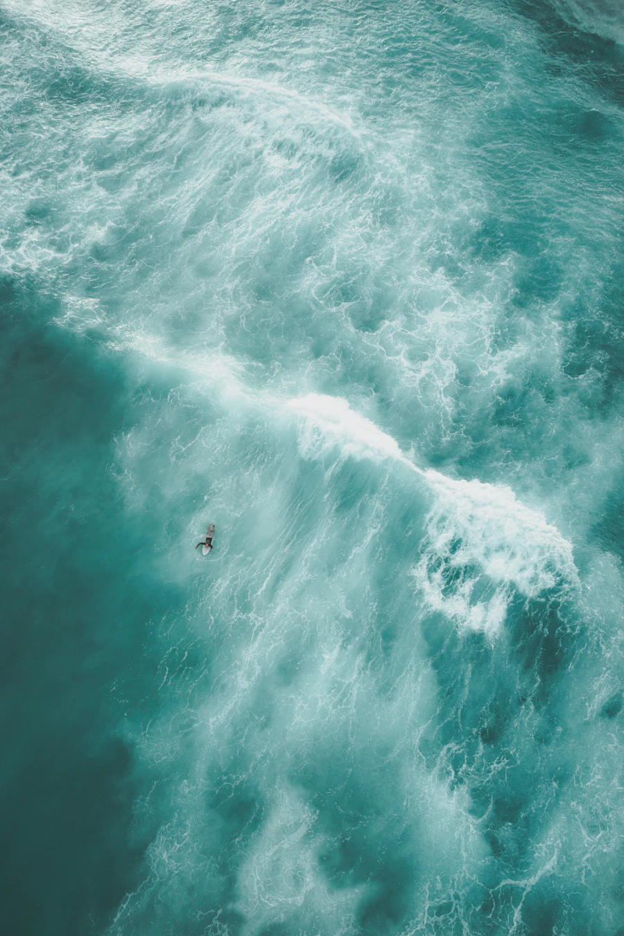A lone surfer drifts beside a breaking turquoise wave seen from above. Foamy white water spreads across the ocean surface.