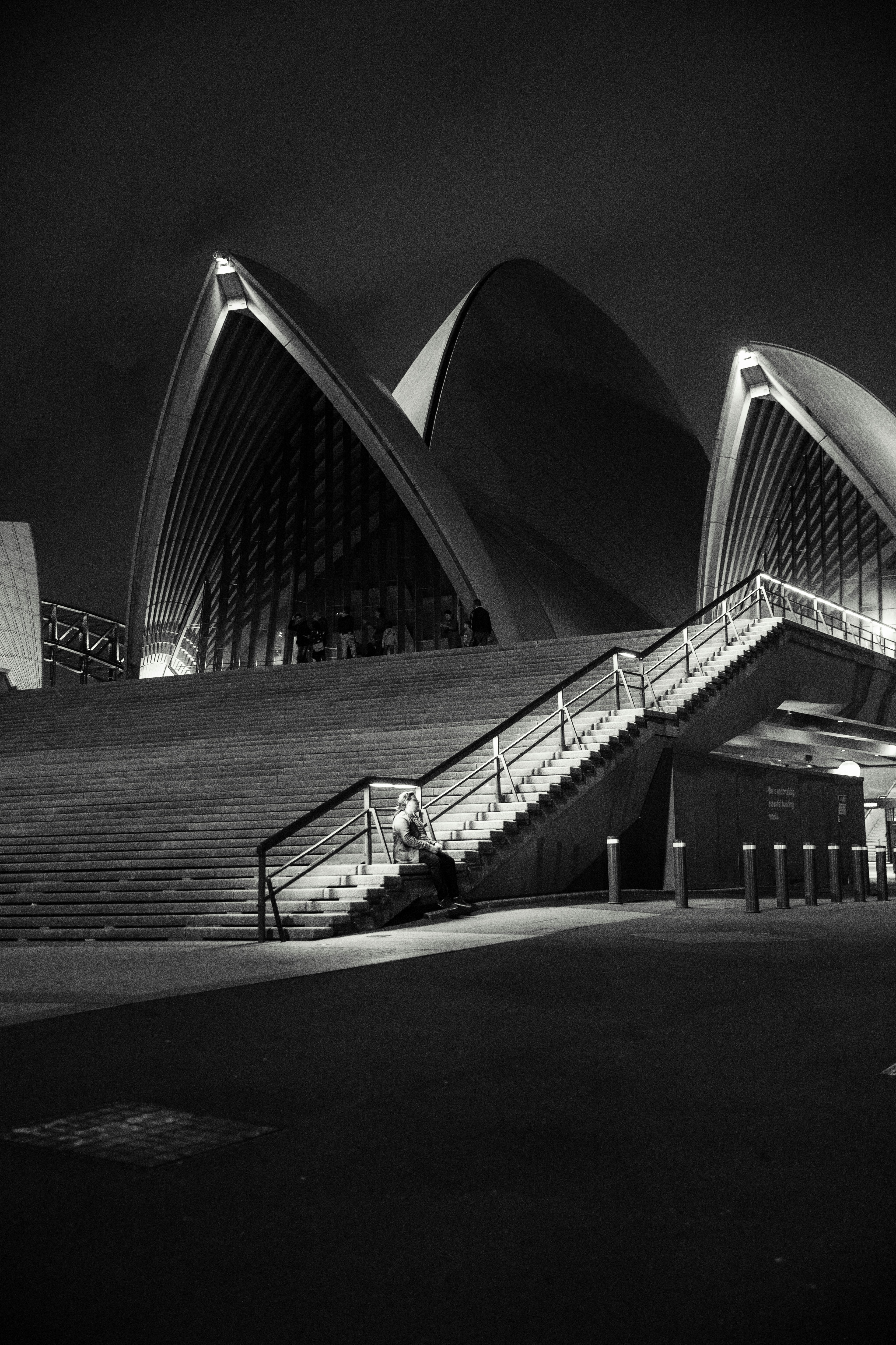 Striking black and white depiction of the Sydney Opera House with dramatic lighting and angular forms. A lone figure rests on the stairs, adding a sense of scale and solitude.