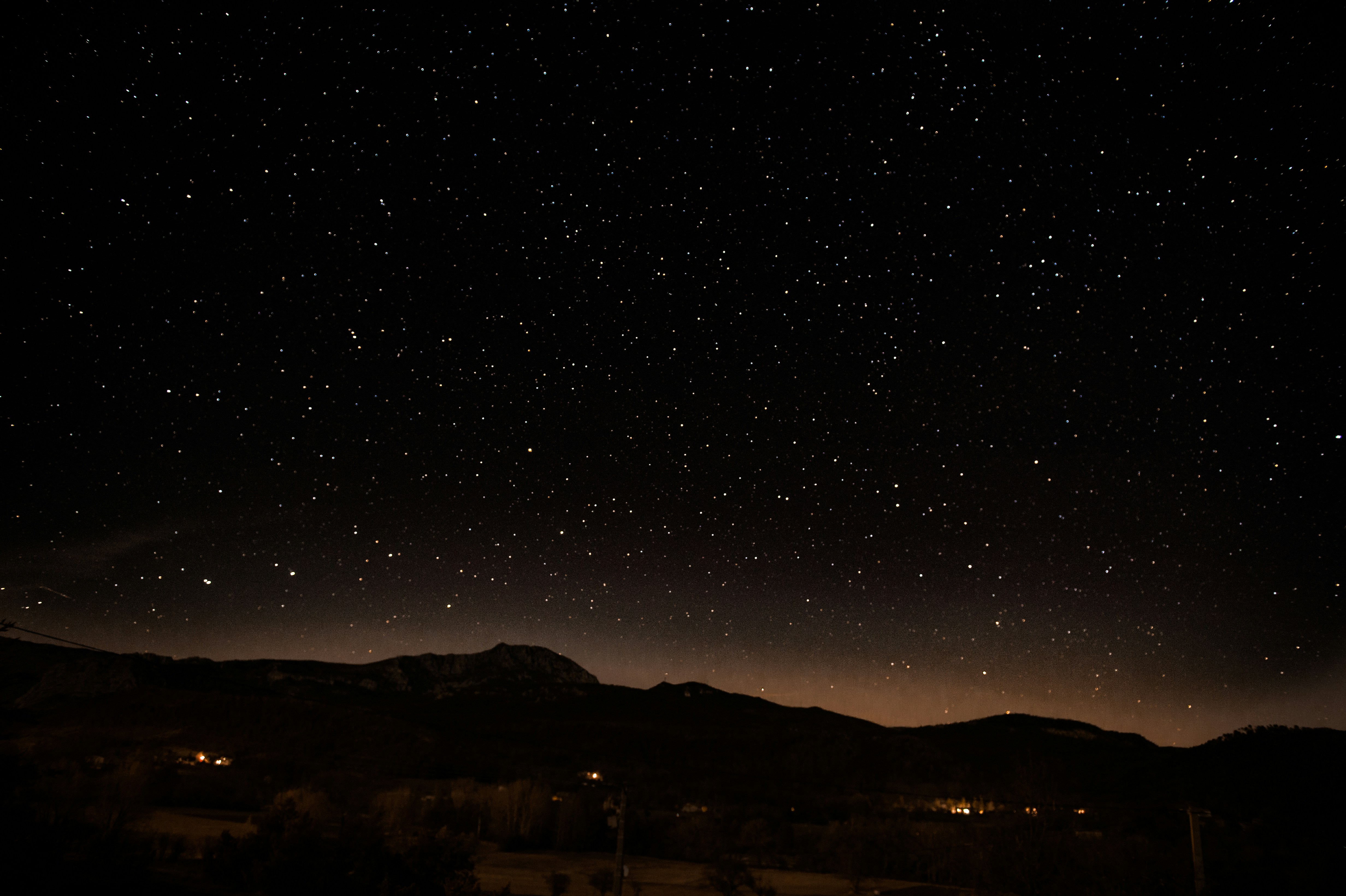 Starry night sky above a silhouette of distant mountains with scattered lights on the horizon.