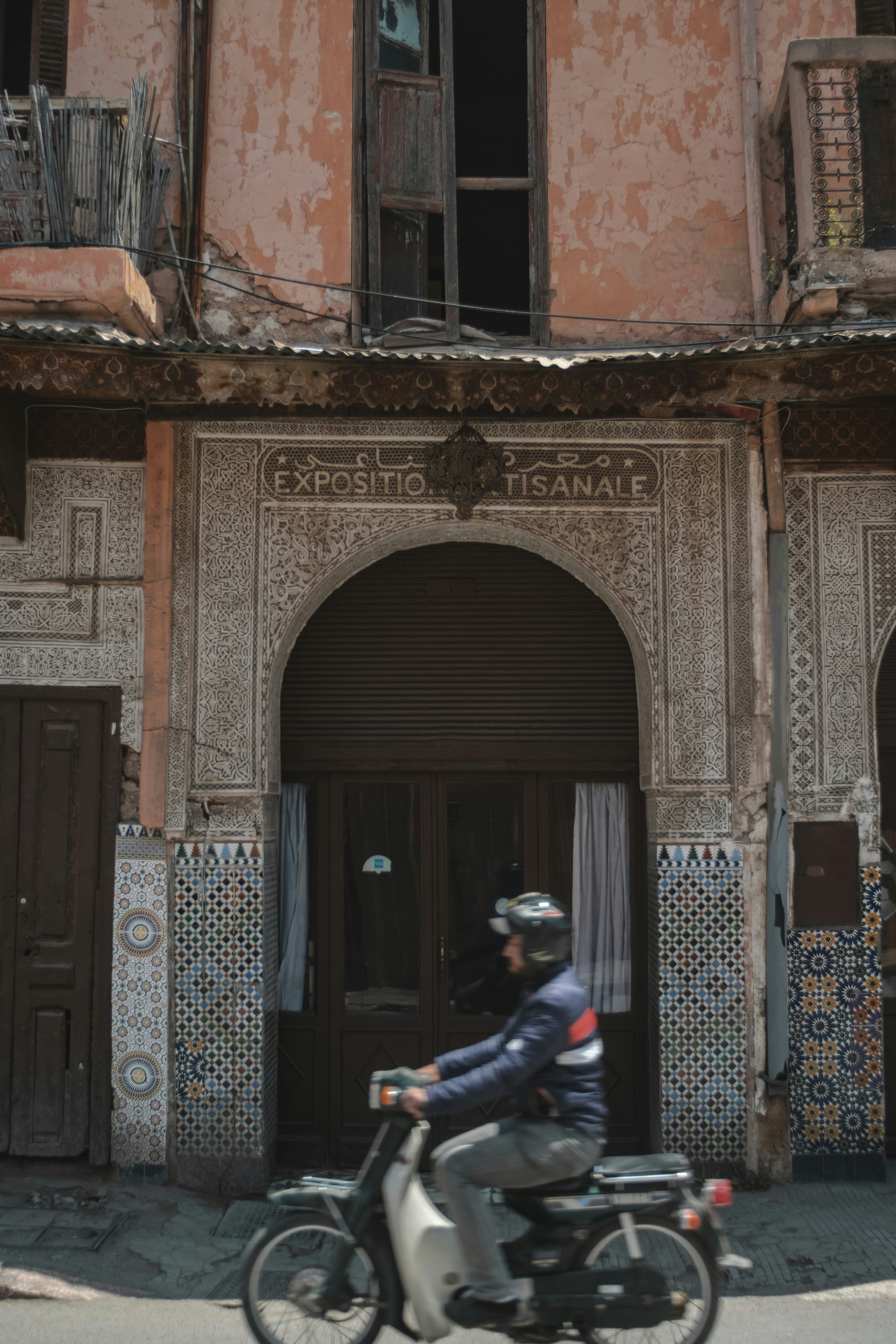 Motorcyclist riding past a traditional artisan shop with intricate tile work and weathered architecture. The scene reflects local craftsmanship and vibrant street life.