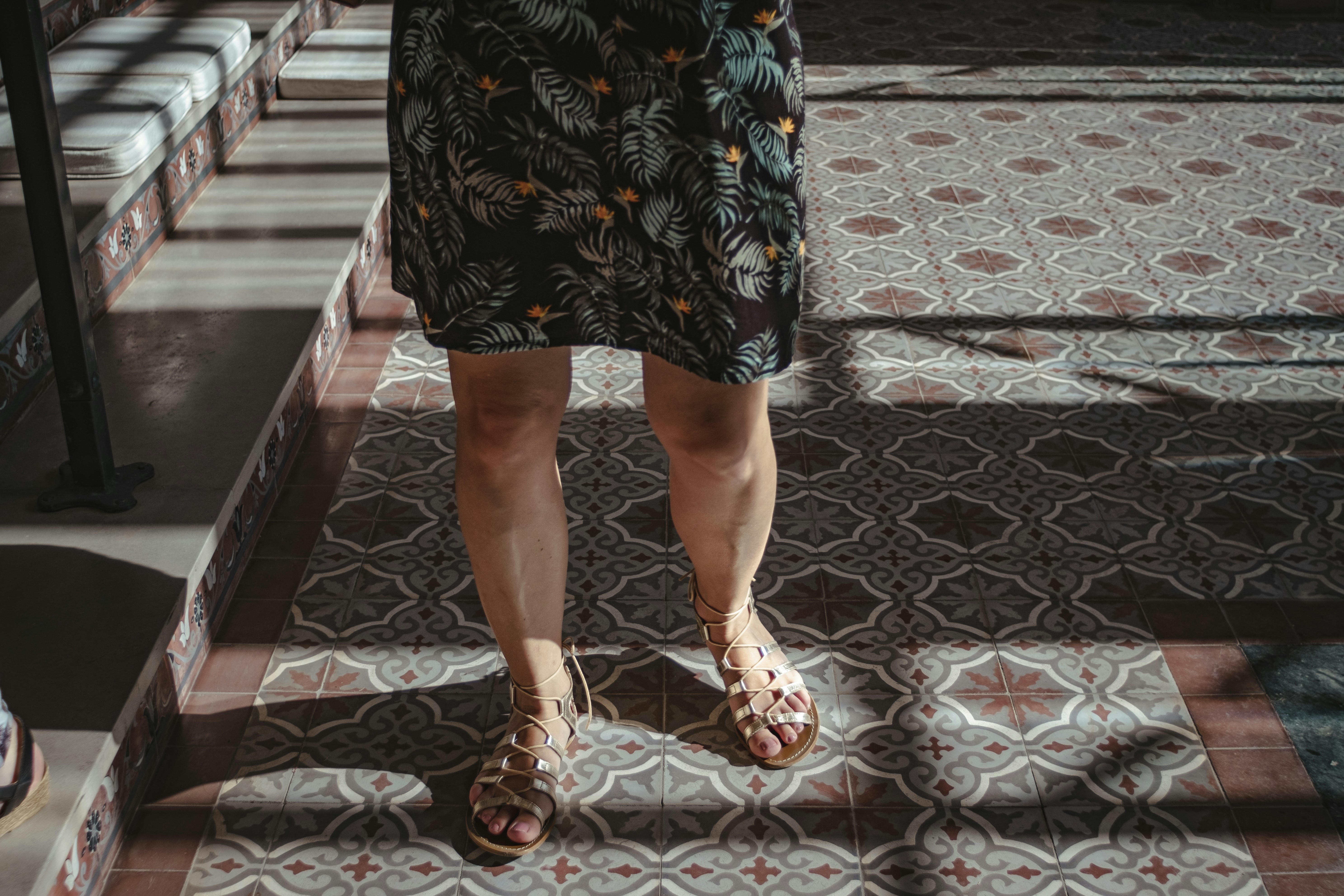 A woman’s legs adorned in a floral dress stand on intricately patterned tiles, with sunlight casting playful shadows around her feet.