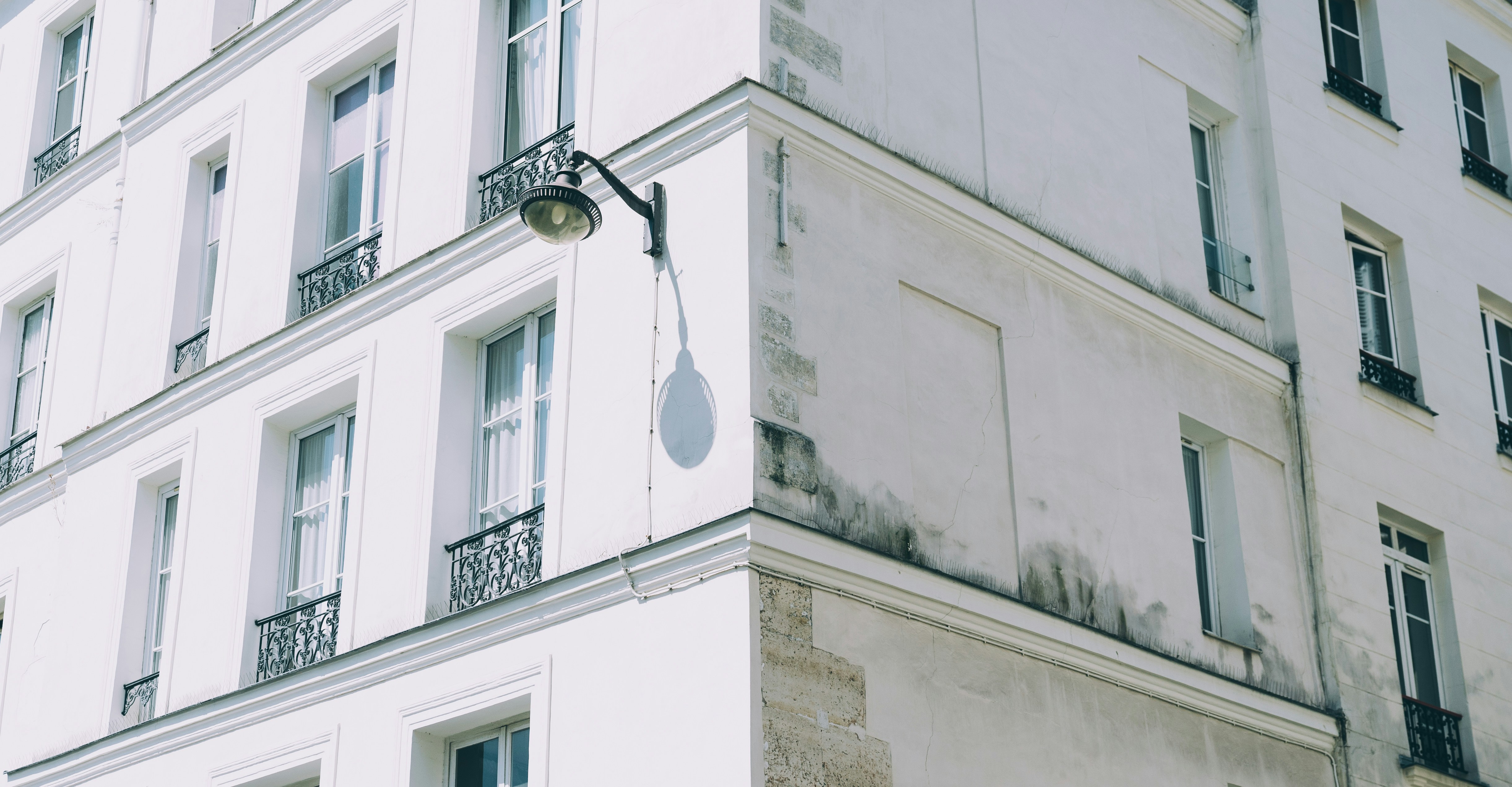 A corner of a white building showcasing intricate window details and a vintage street lamp casting a shadow.