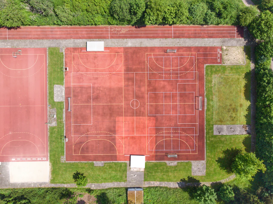 Top-down view of red outdoor basketball and tennis courts bordered by green trees and grass. White court lines and small shelters stand out against the worn playing surfaces.