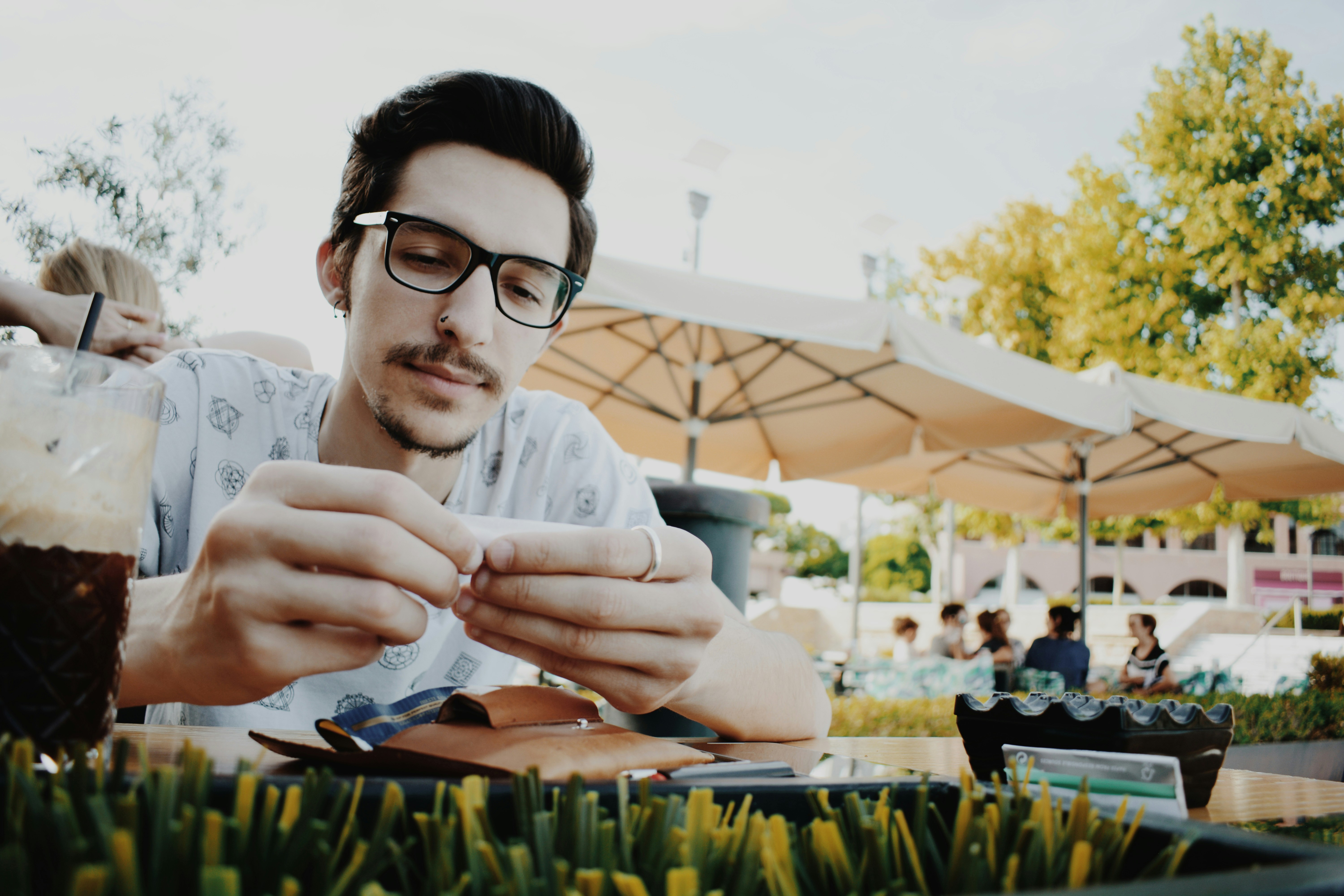 Young man engaged in a focused activity at an outdoor café, surrounded by greenery and casual conversation. Warm sunlight enhances the relaxed atmosphere.