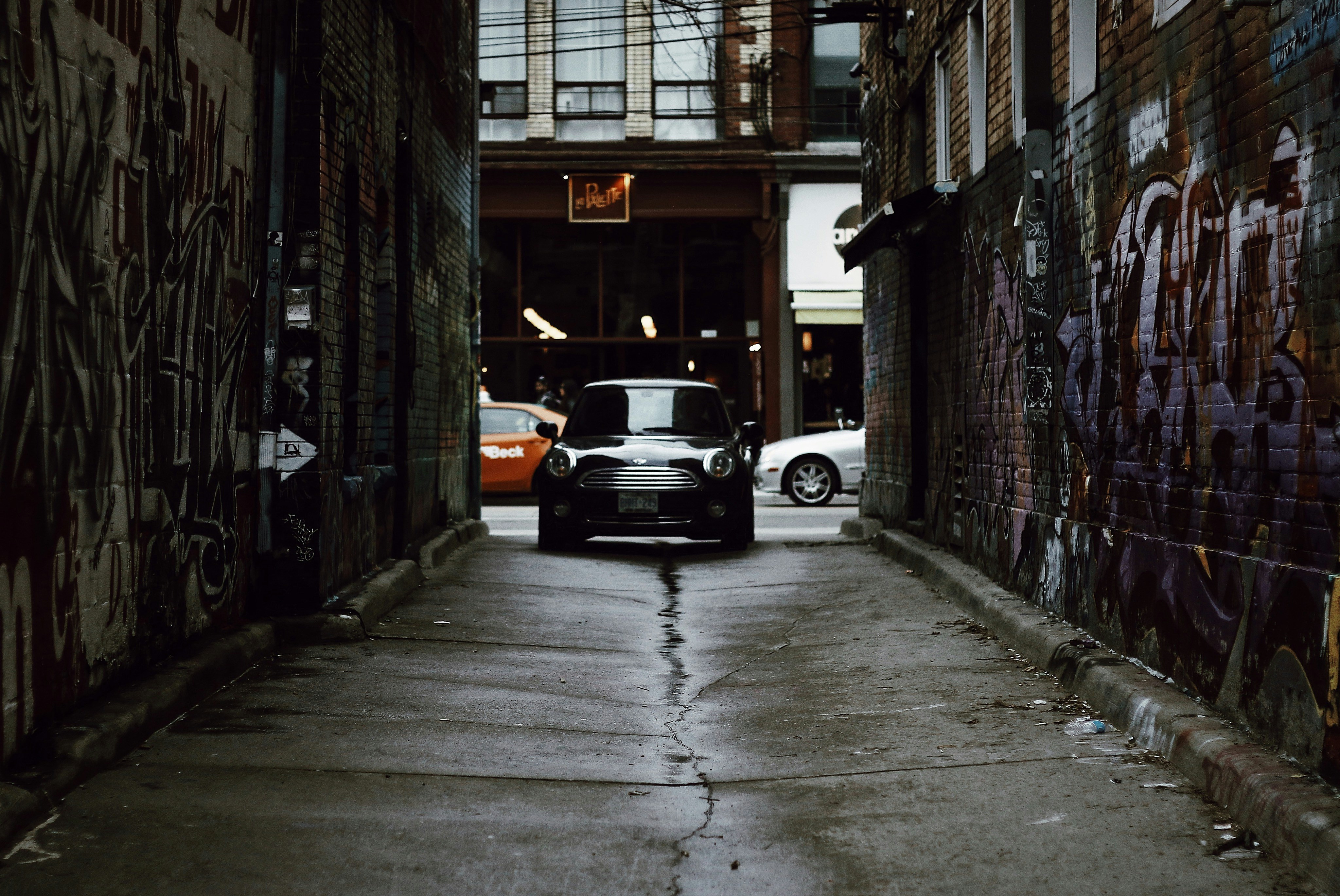 Graffiti-covered alley with a car parked at the end, framed by urban architecture.