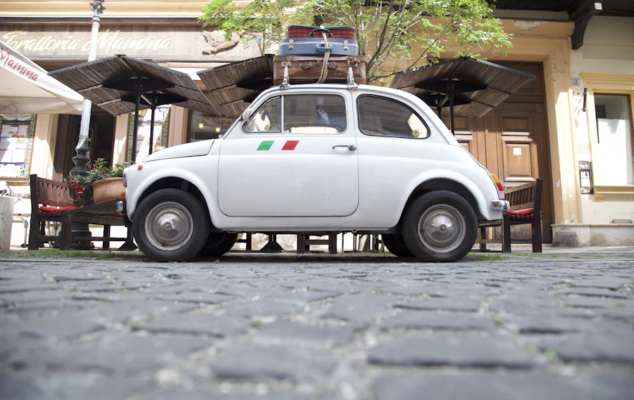 A white vintage Fiat 500 with Italian flag decals is parked in front of a café or trattoria. Cobblestone pavement and outdoor seating frame the scene.