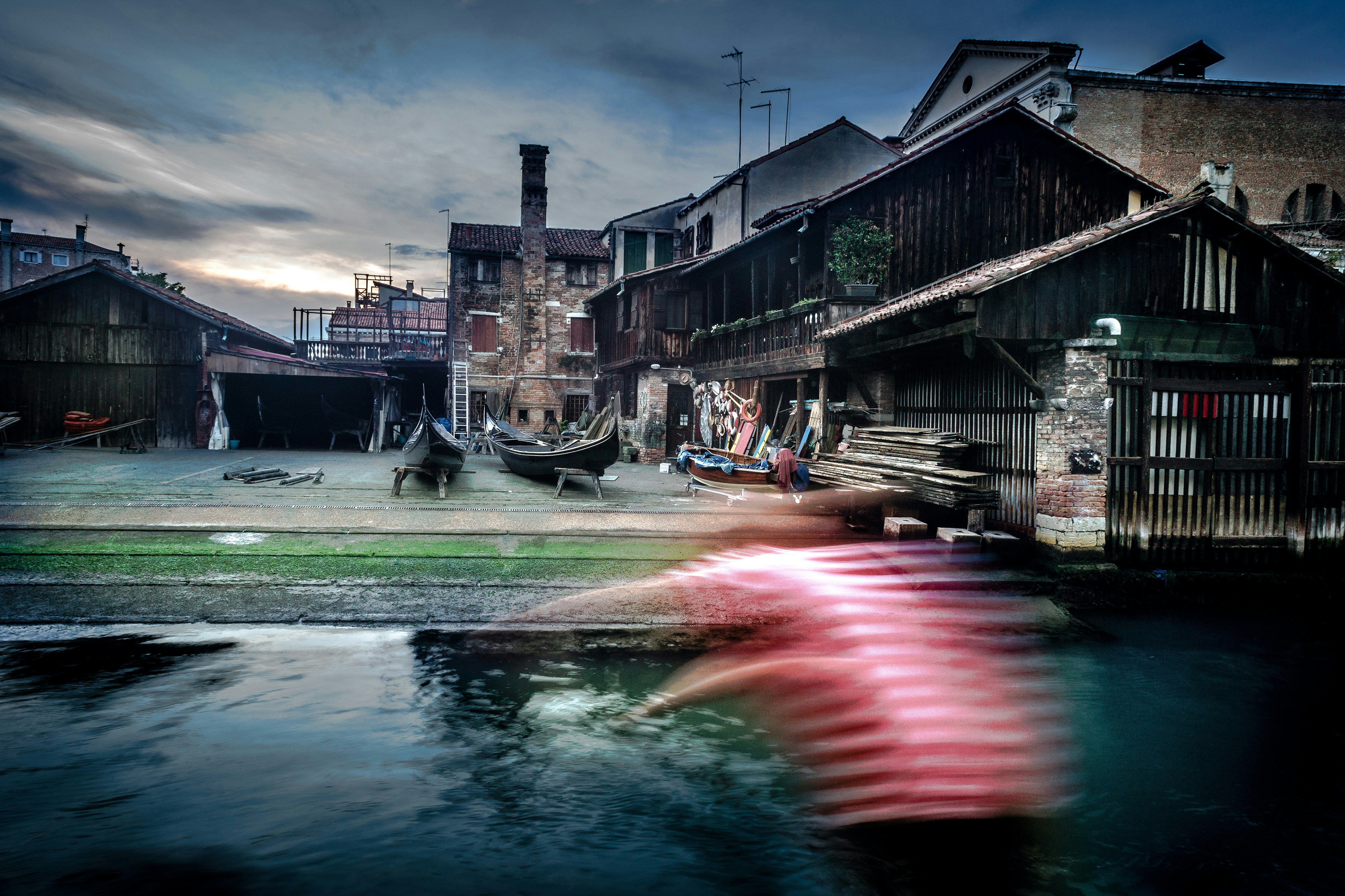 Blurred motion of a passing boat along a canal with rustic warehouses and boats at dusk; foreground water reflects the scene while a long exposure softens the movement.