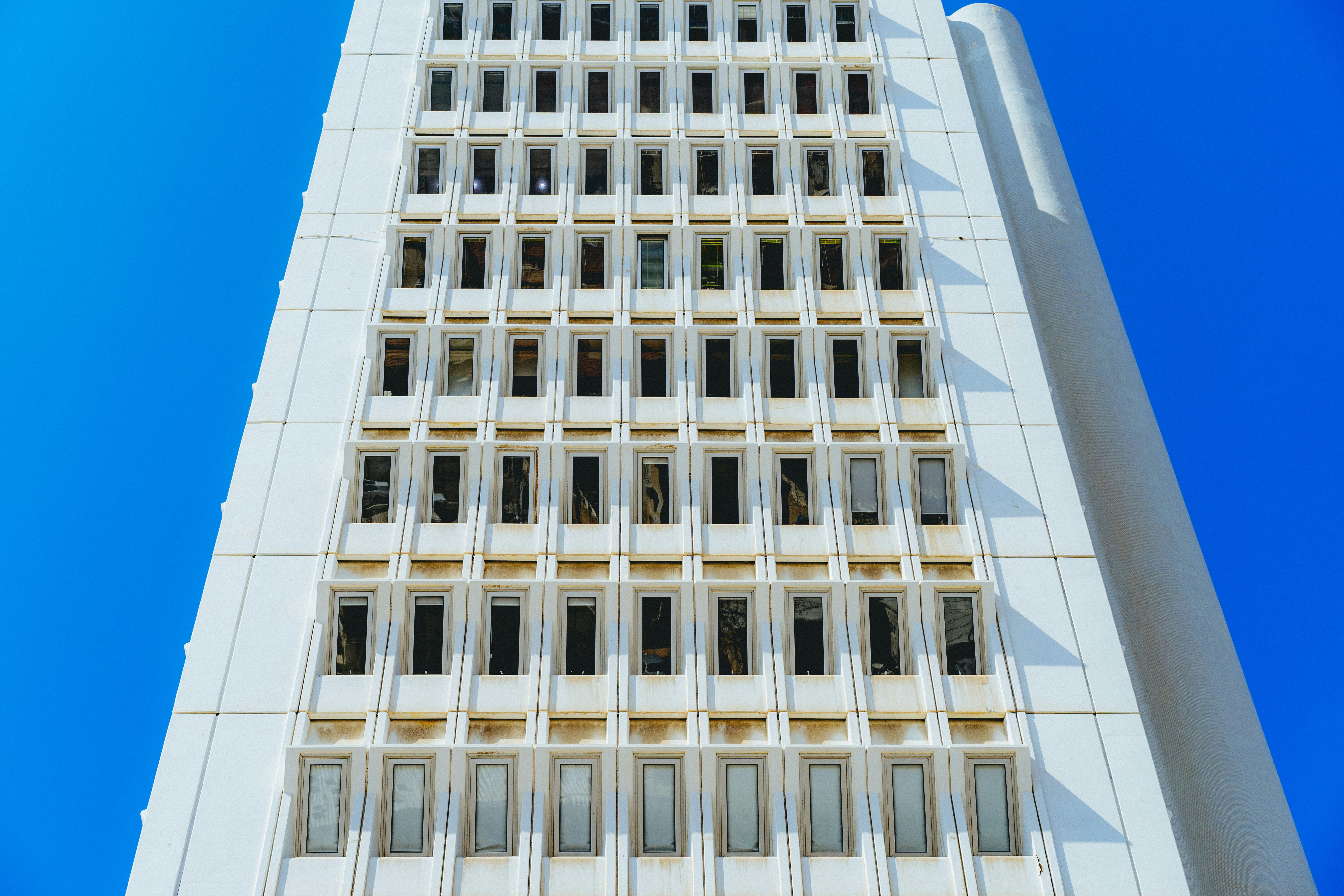A high-rise building showcasing a rhythmic pattern of windows against a clear blue sky. The stark white facade emphasizes modern architectural design.