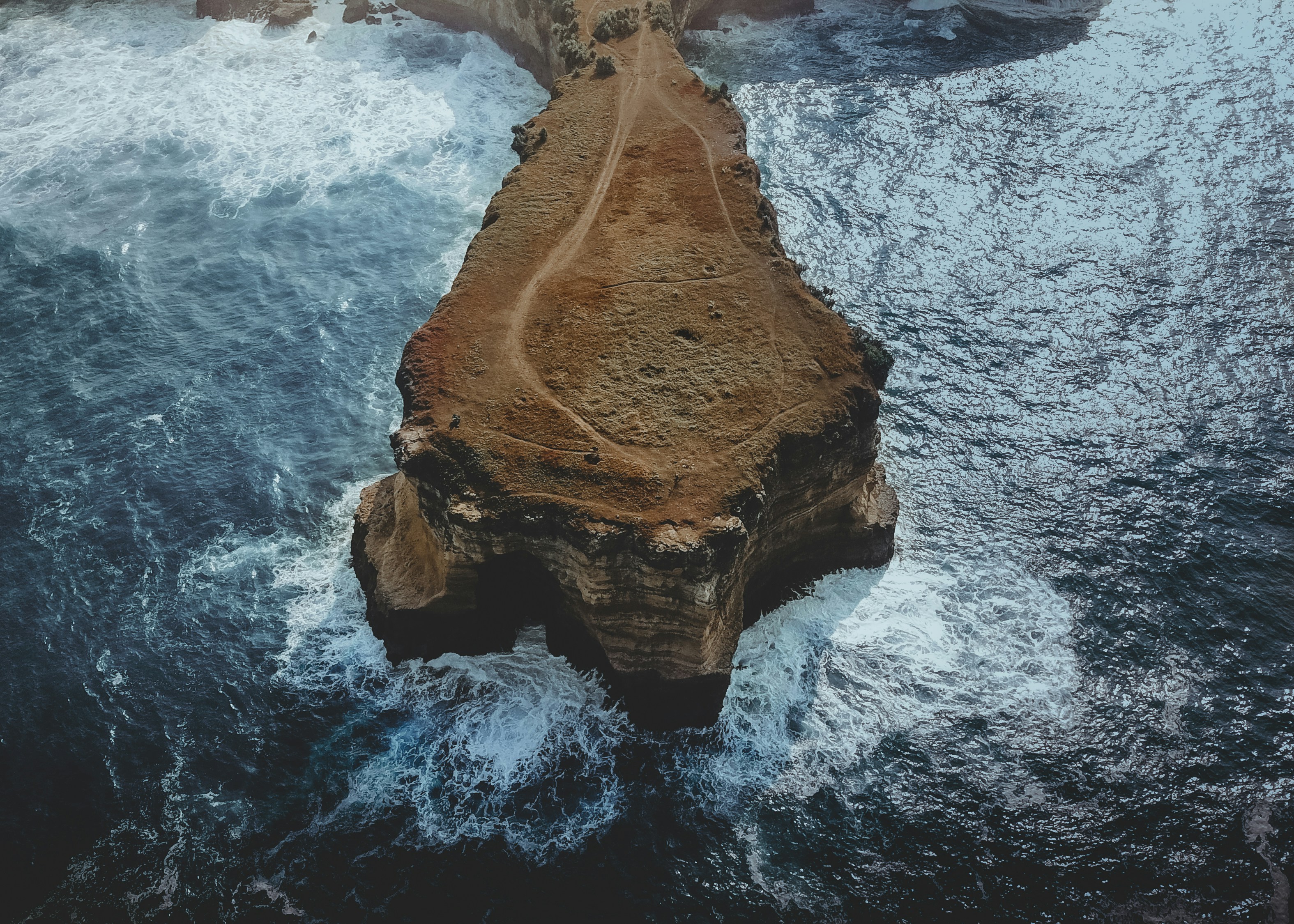 Aerial view of a rugged coastal landform surrounded by turbulent ocean waves, highlighting the stark contrast between land and sea.