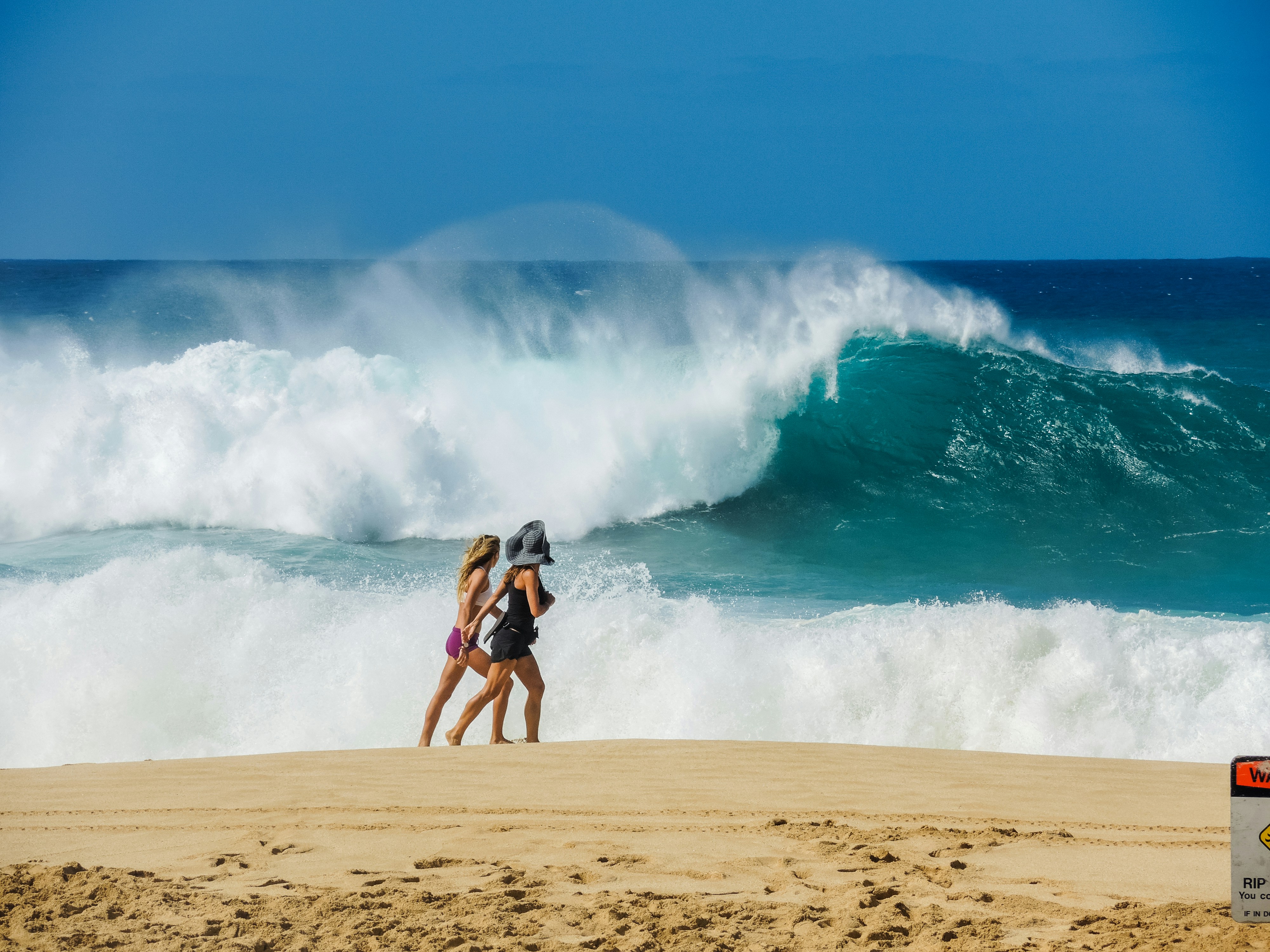 Two women walk along a sandy beach with powerful waves crashing in the background.