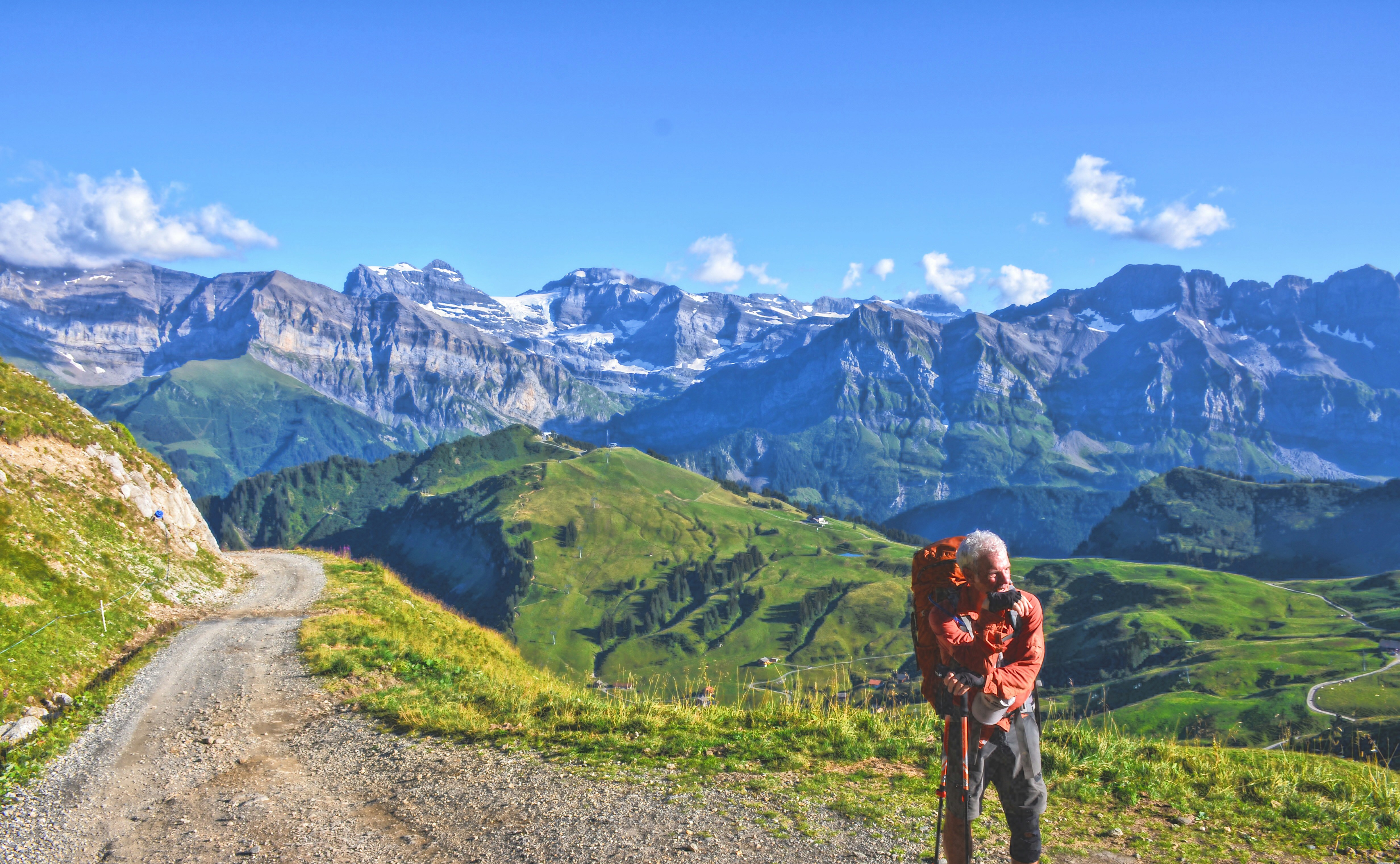 Hiker pauses on a winding gravel path, surrounded by towering mountains and lush green valleys under a clear blue sky.