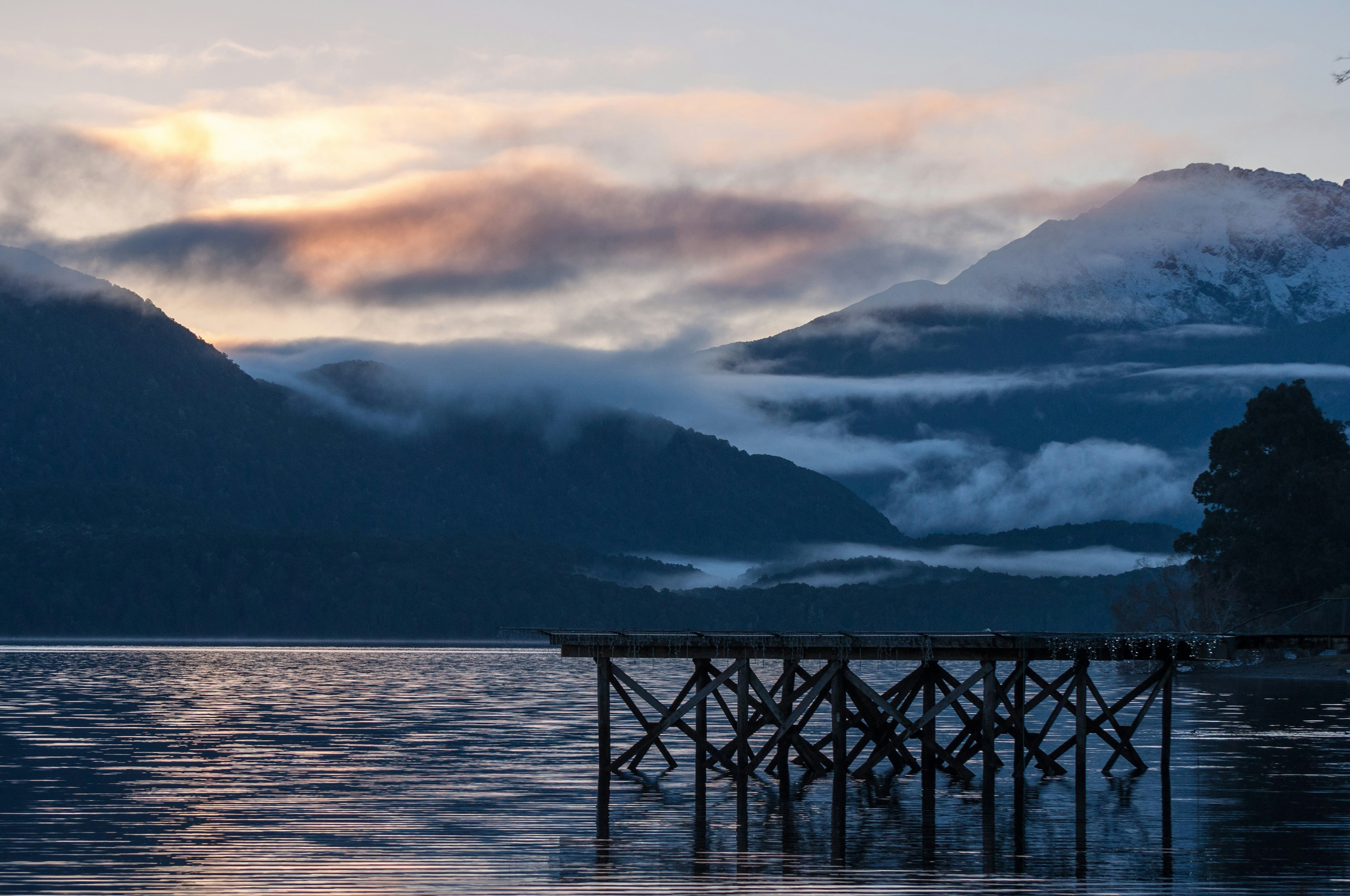 Wooden pier extends into a tranquil lake with misty mountains and a colorful sunset sky.