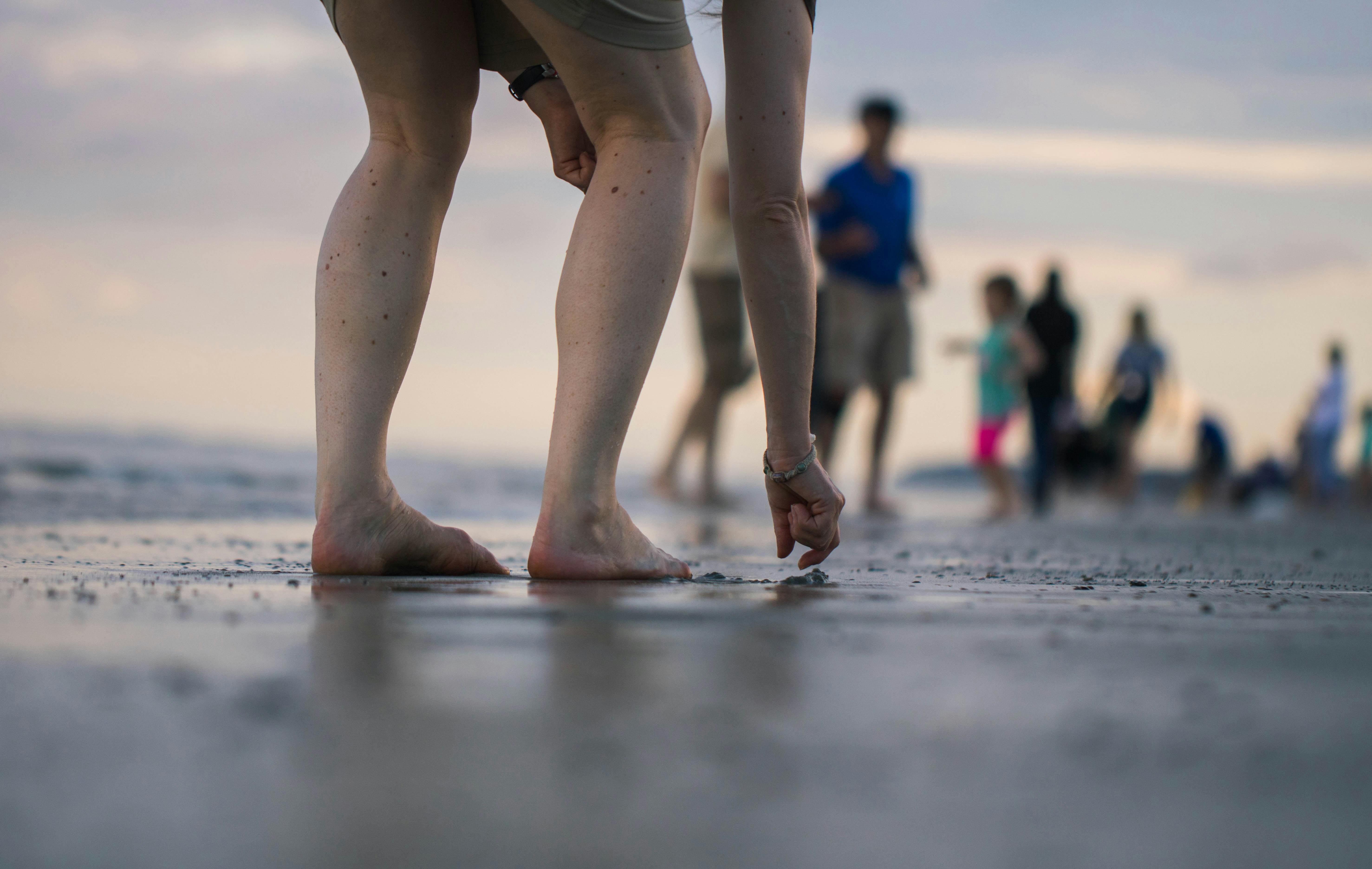 Person crouching on wet sand near a shoreline at sunset, with blurred figures in the background.
