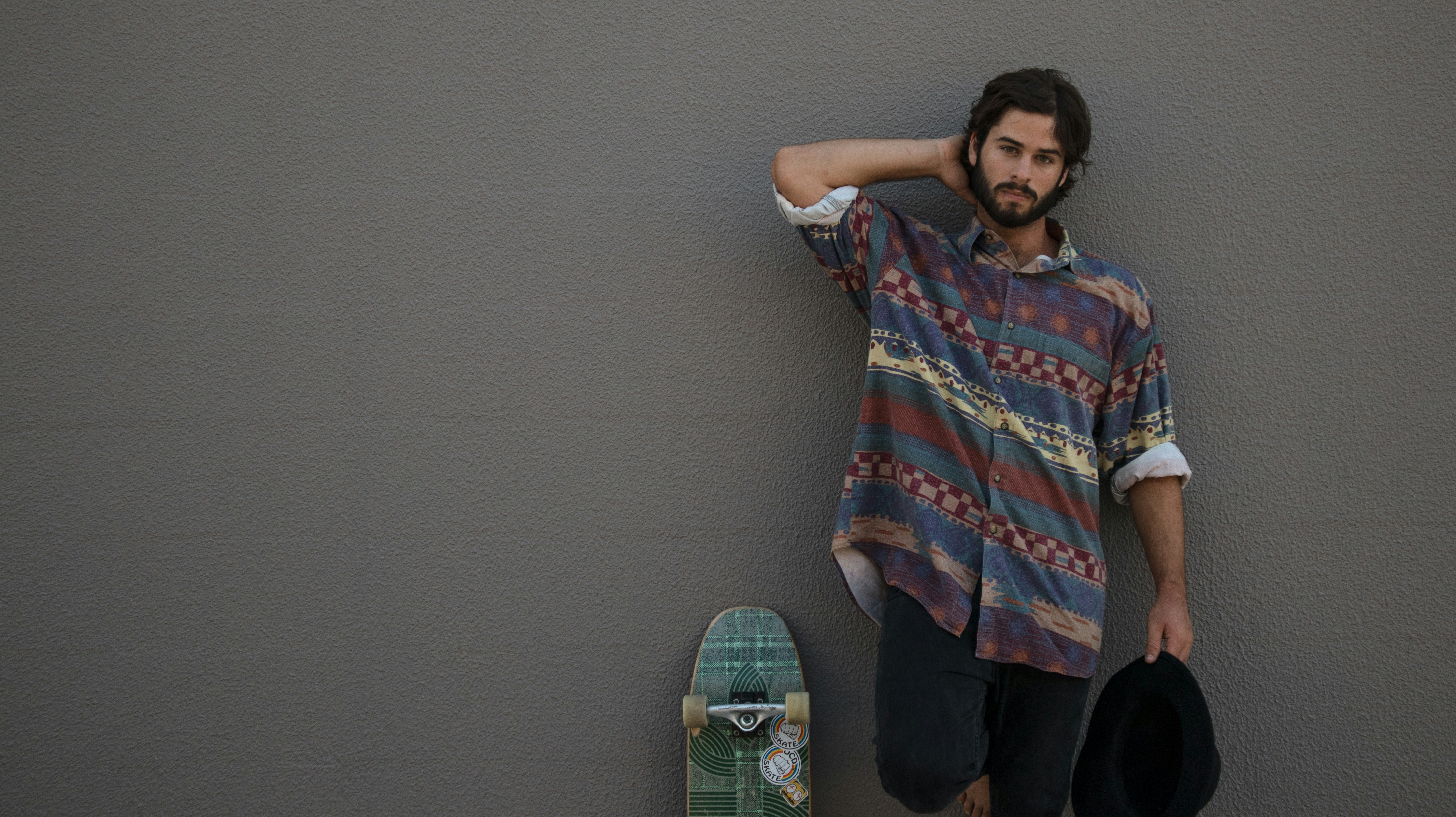 Man in patterned shirt and shorts leaning against a wall with a skateboard.
