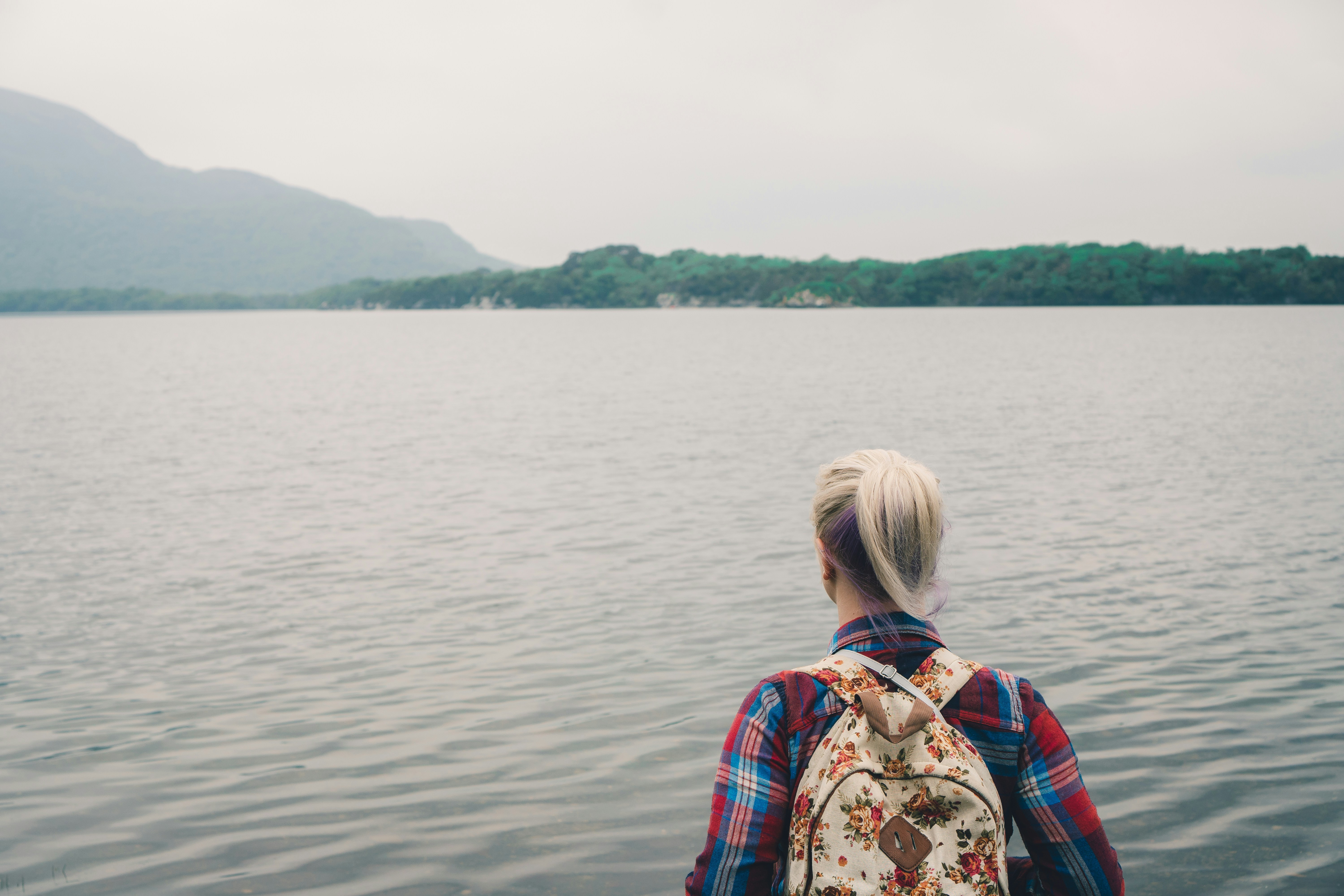 Person with a floral backpack gazing across a tranquil lake towards distant green hills.