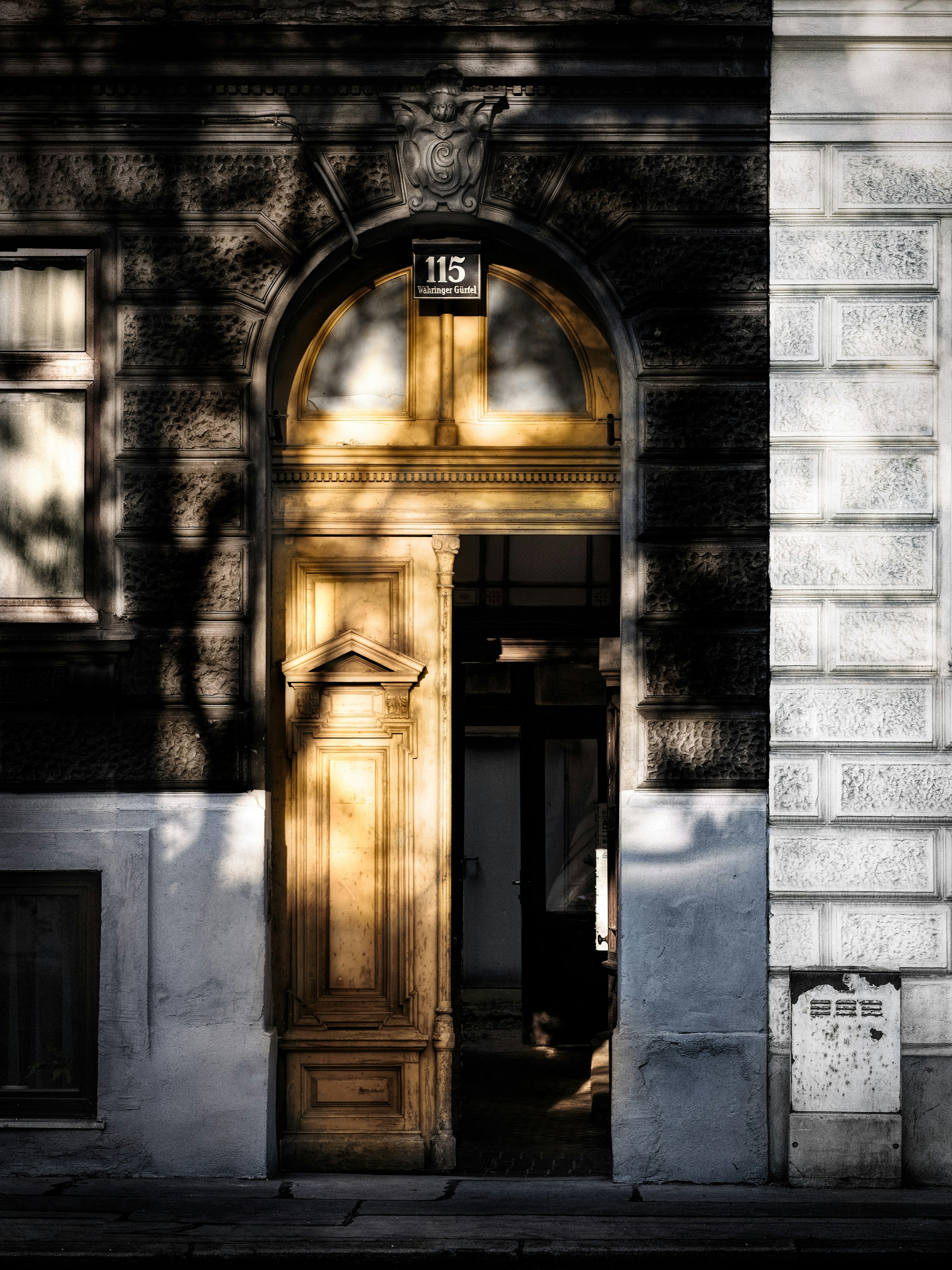 Intricately carved wooden door framed by contrasting shadows, revealing a glimpse into a historic interior. The door is marked with number 115.