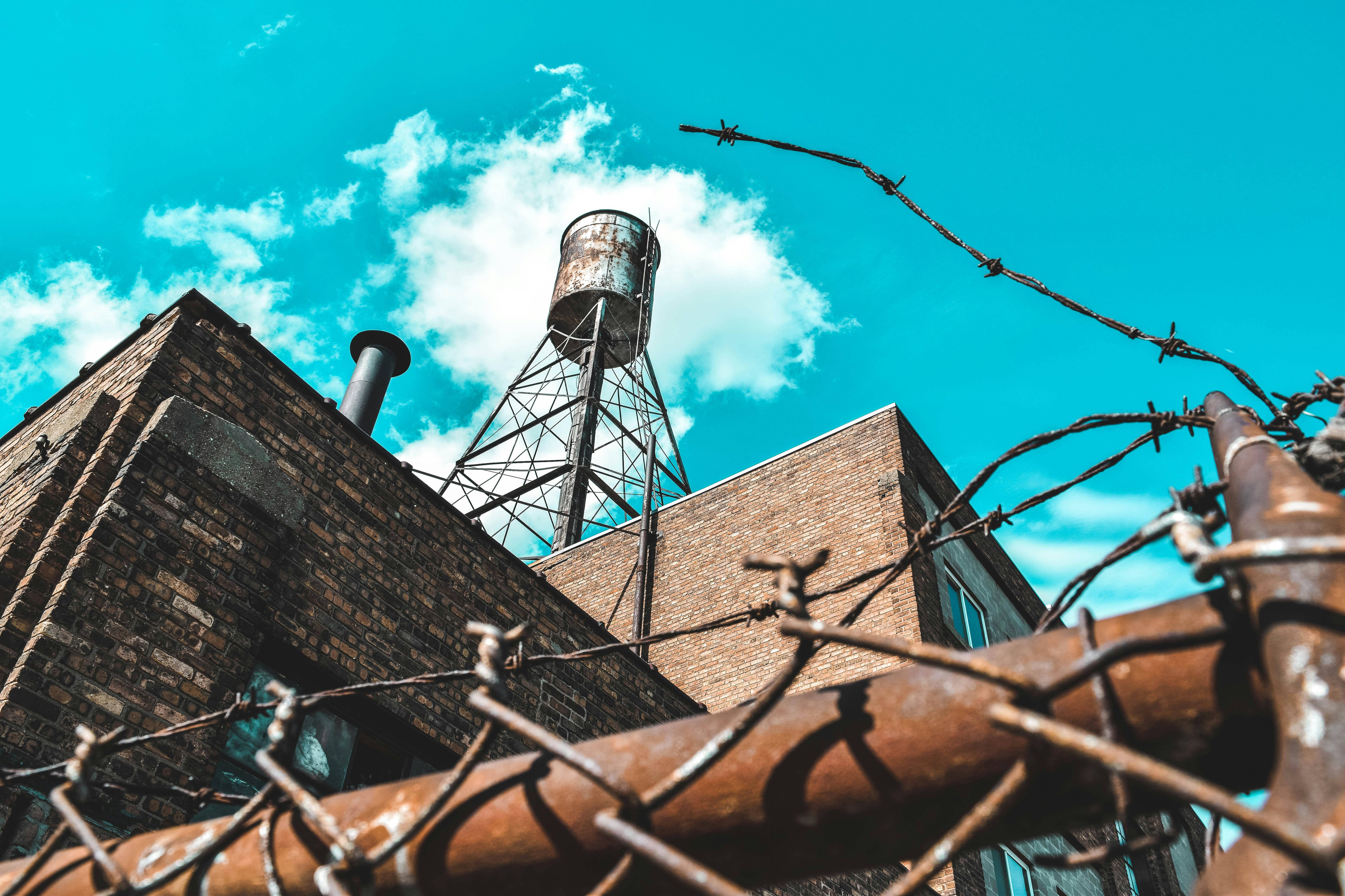 Low-angle view of a brick building and water tower framed by barbed wire against a vibrant blue sky.